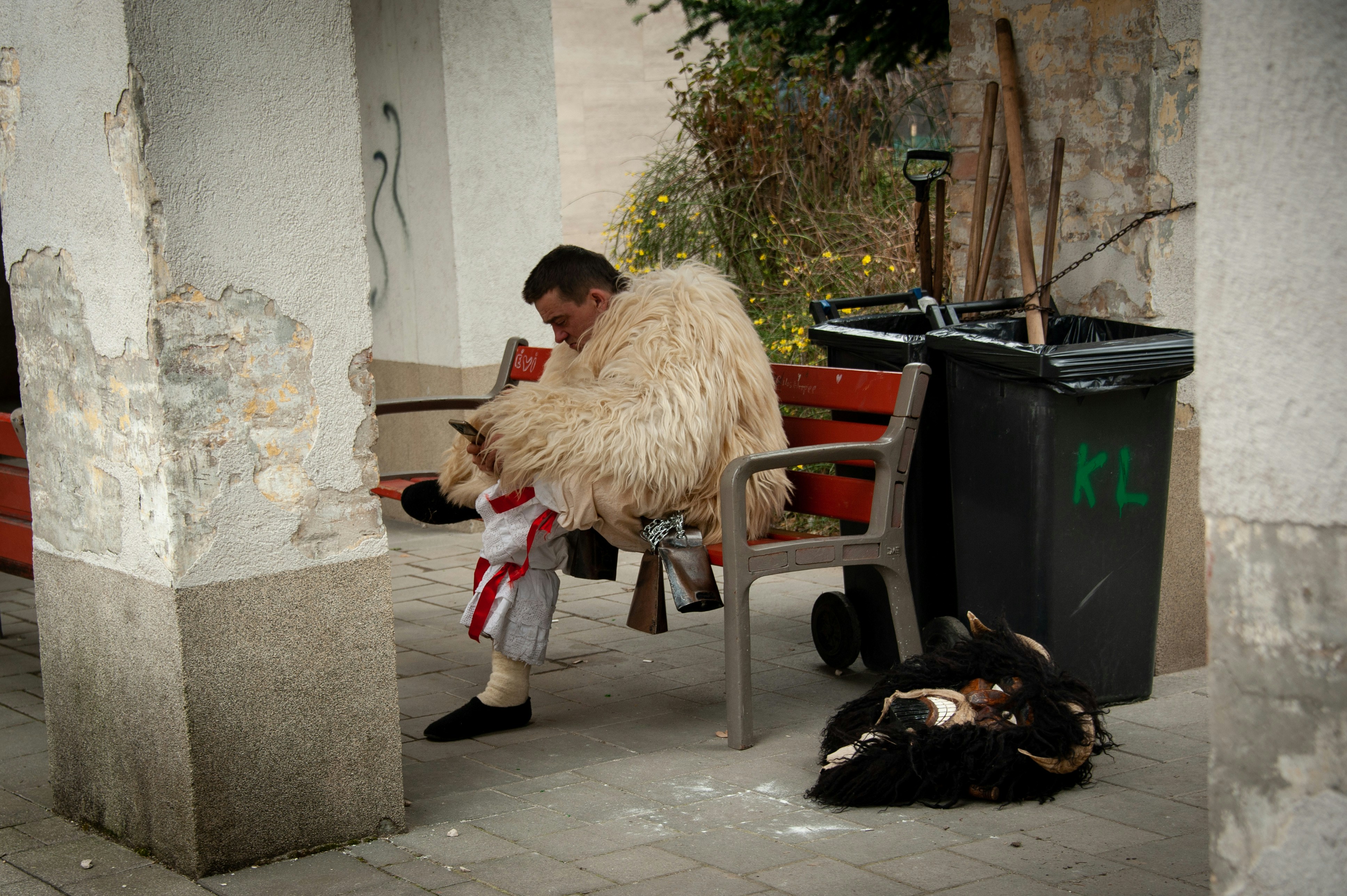 A performer in traditional attire sits on a bench, absorbed in his phone, while a discarded mask lies nearby. The setting reflects a blend of cultural heritage and modern life.