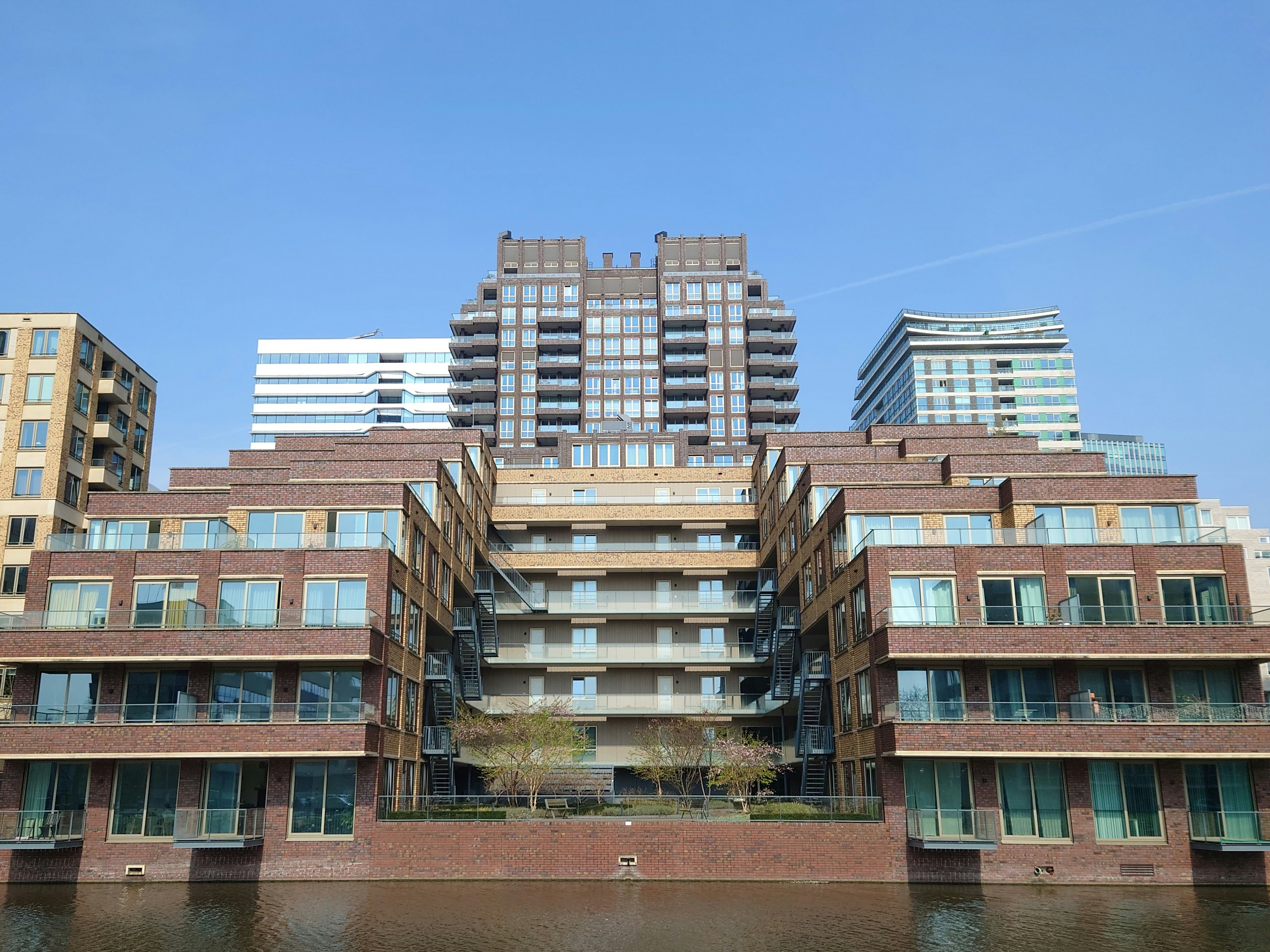 Symmetrical, tiered residential and office buildings beside a calm waterway under a clear blue sky.