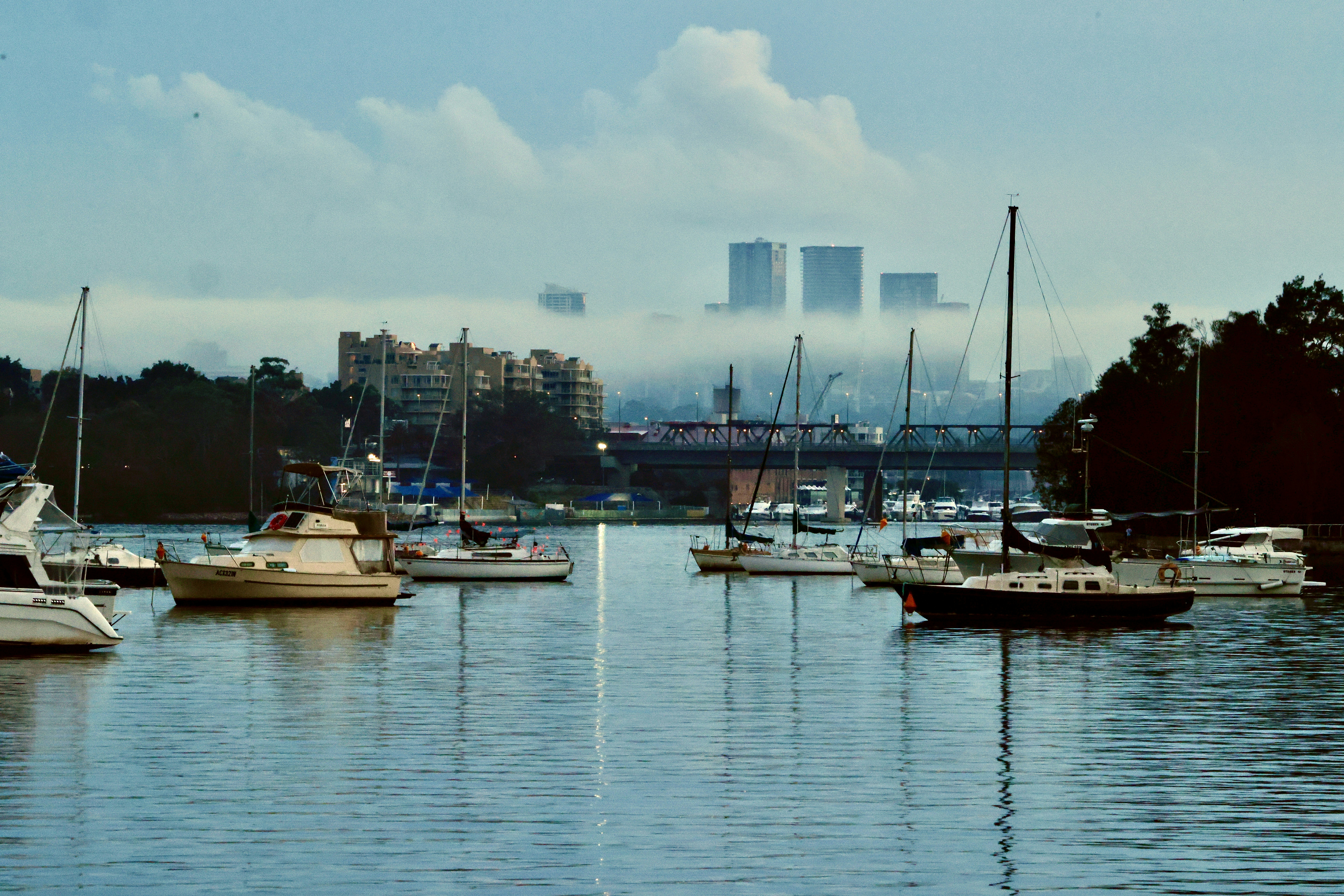 Sailboats rest on calm waters with a city skyline visible through morning mist.