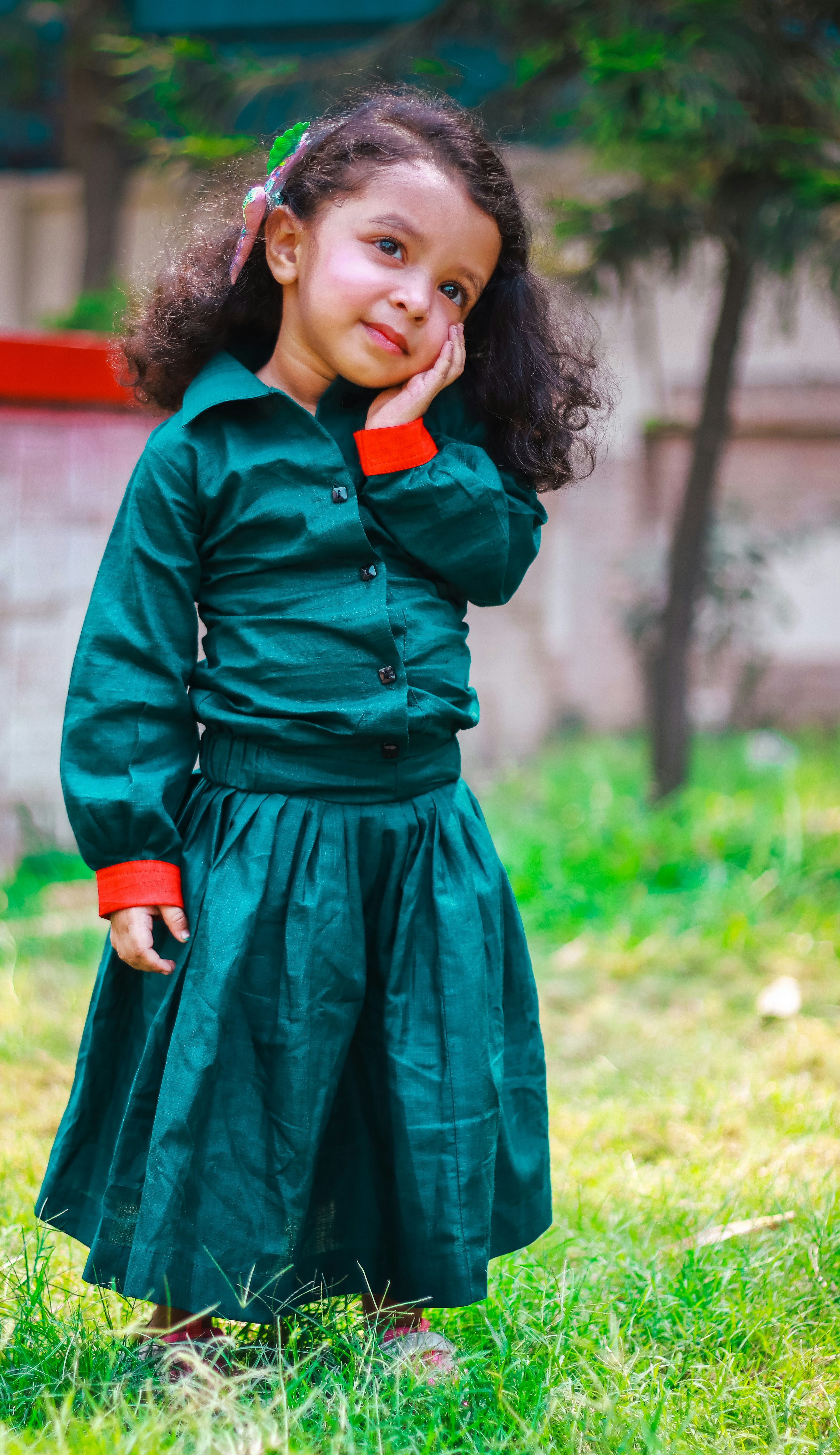 Young girl in a green outfit with a playful pose, surrounded by lush greenery. Her expression conveys a sense of curiosity and charm.