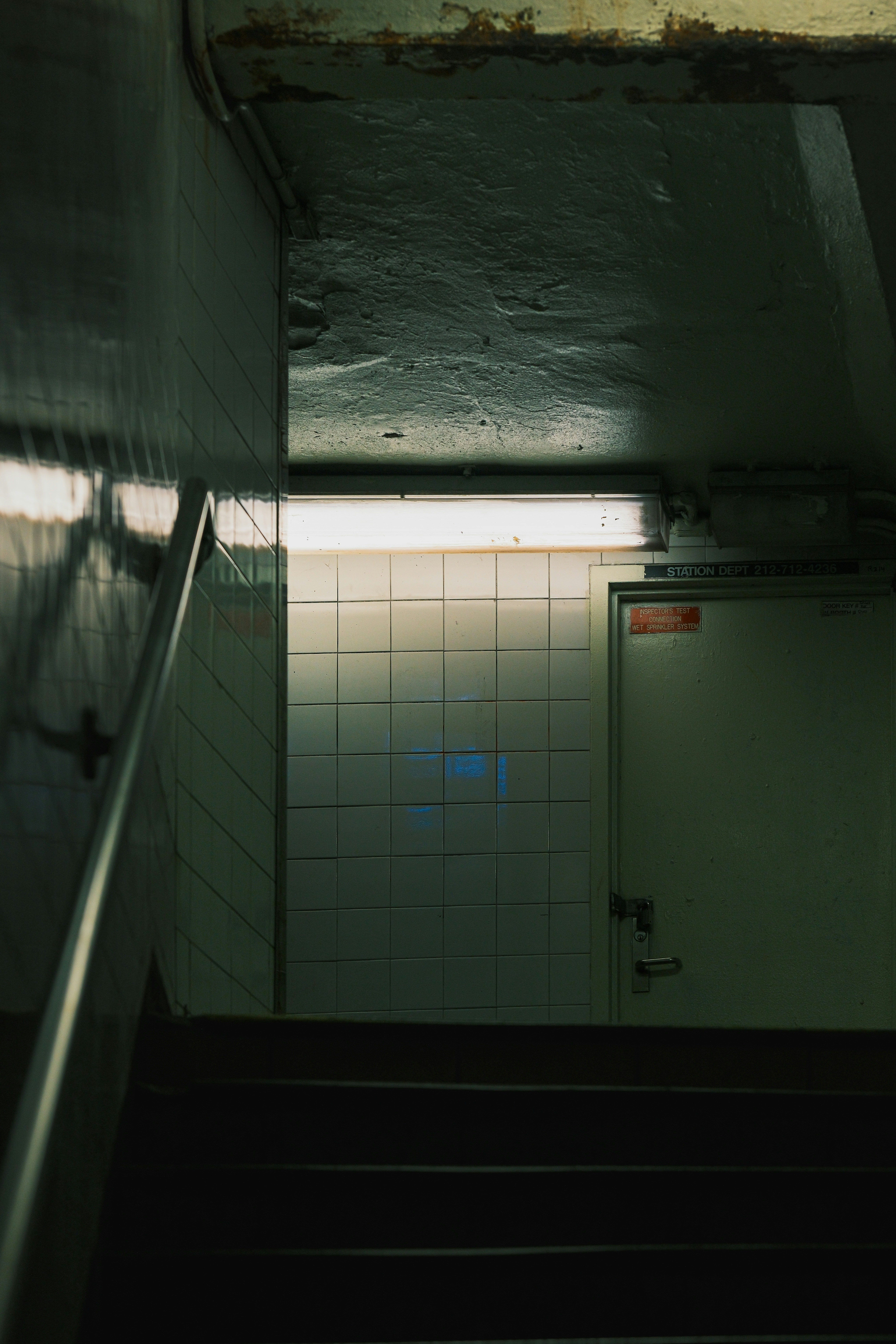 Dimly lit subway staircase leading to a door marked with station information. The interplay of light and shadow creates a moody atmosphere.