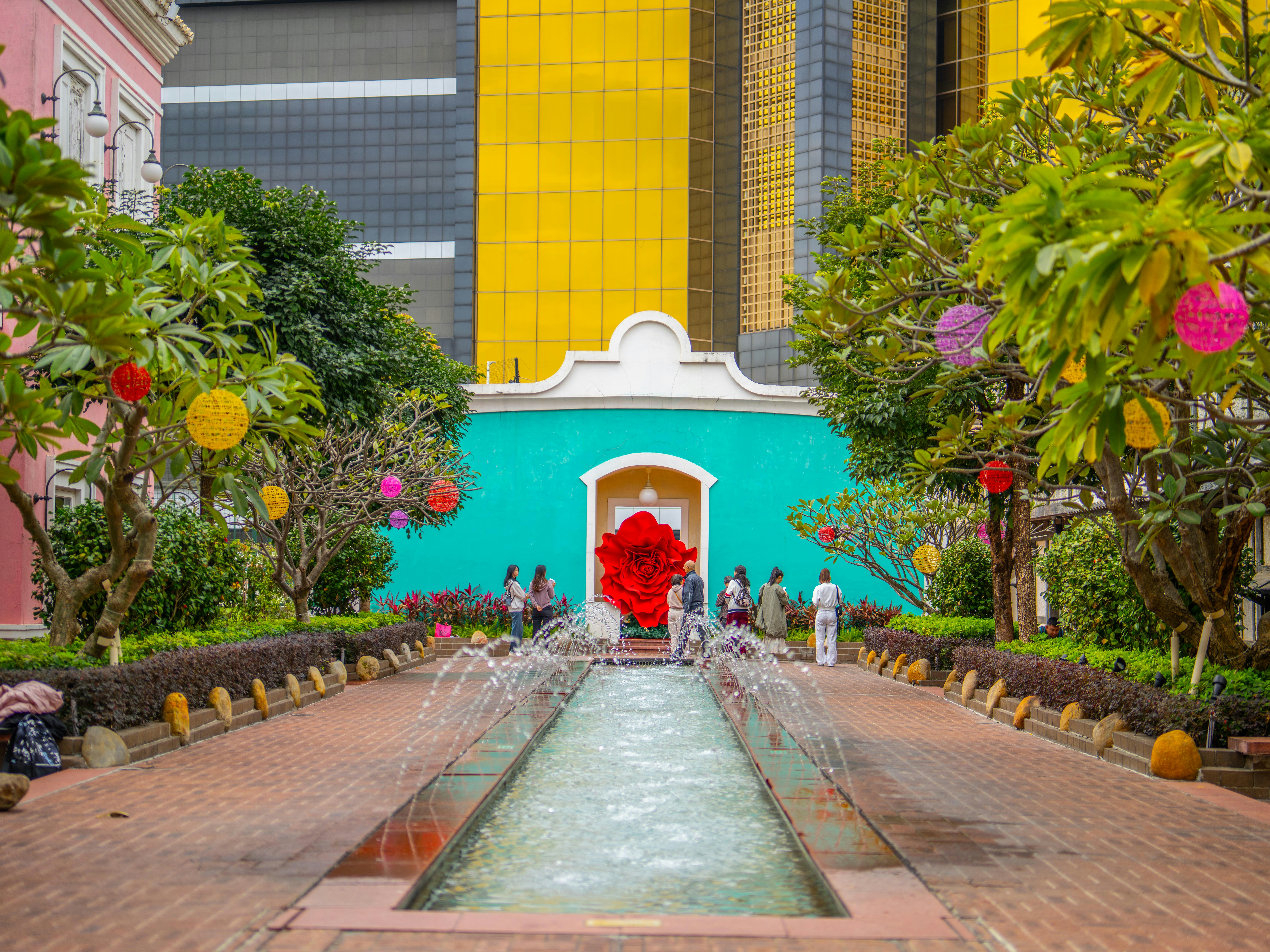 Lush garden path with colorful lanterns leading to a turquoise wall and red door, juxtaposed against a modern cityscape.