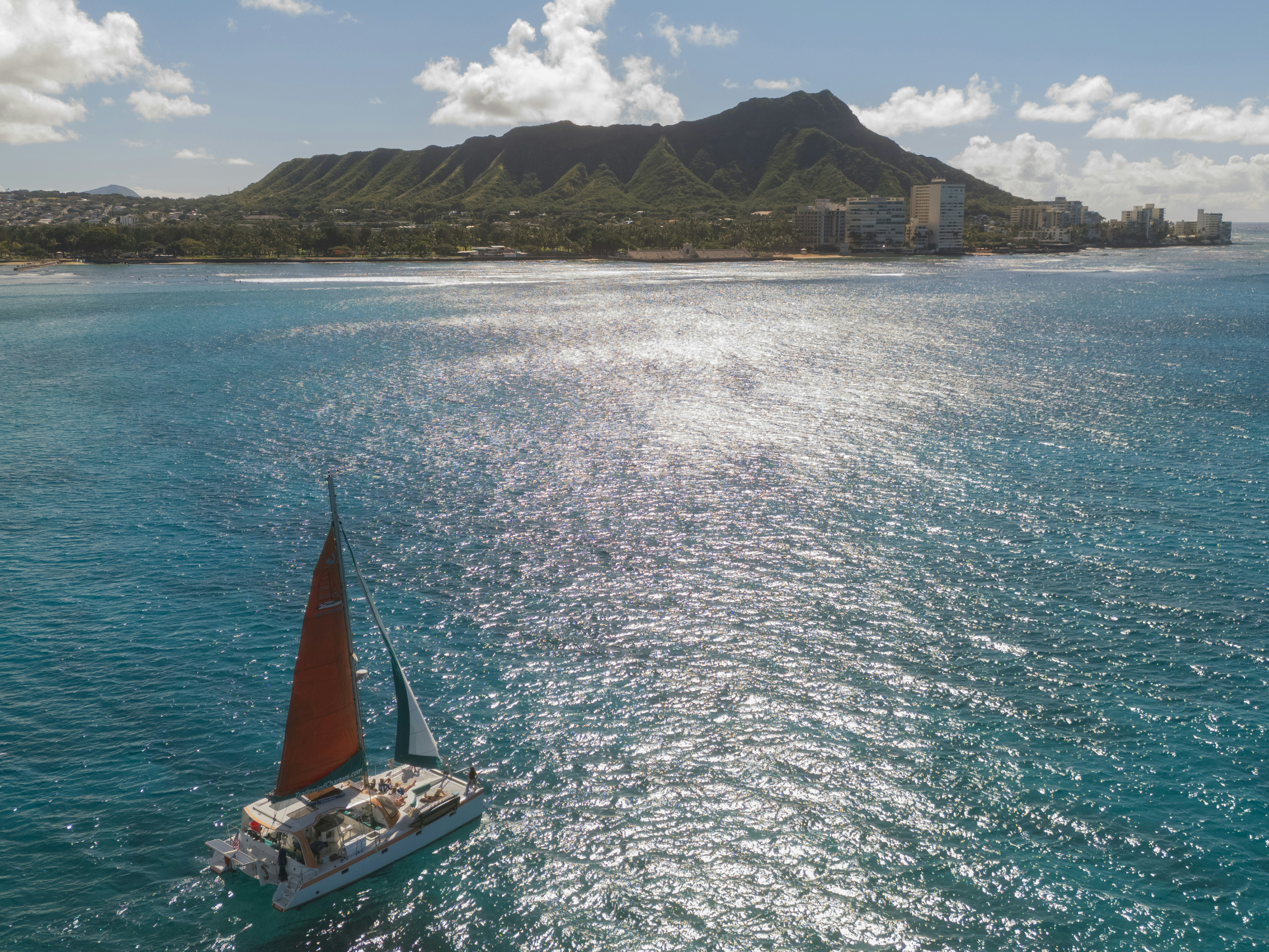 Catamaran with vibrant sail glides across turquoise ocean near Diamond Head under a bright sky.