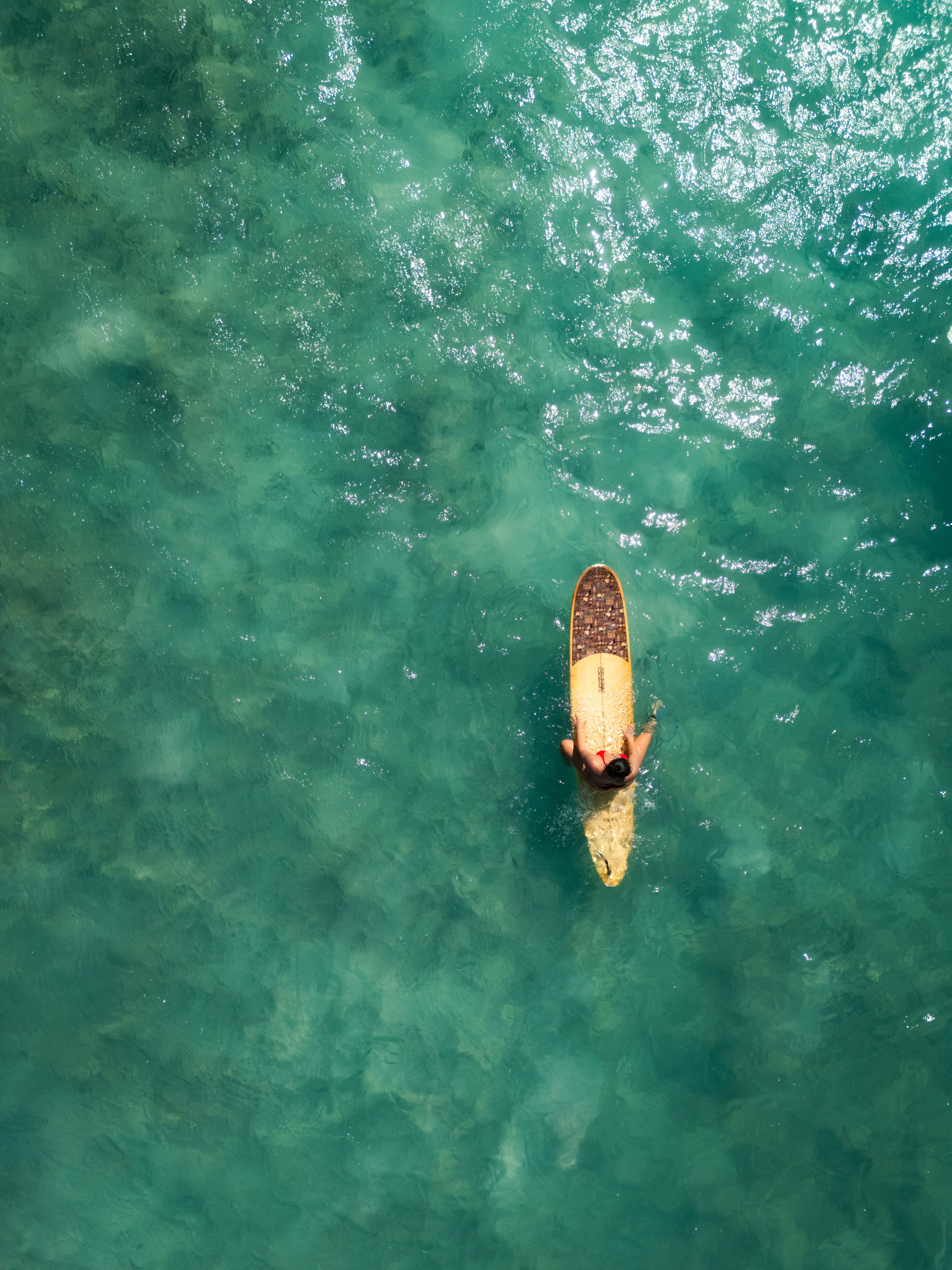 An aerial shot captures the peaceful moment of a lone surfer paddling on a wooden surfboard in stunning turquoise waters. The ocean's clarity reveals subtle textures beneath the surface, while sunlight sparkles across the scene, evoking a sense of calm and adventure. This image embodies the essence of tropical leisure and connection with nature, making it ideal for themes related to travel, surfing culture, or coastal lifestyles.