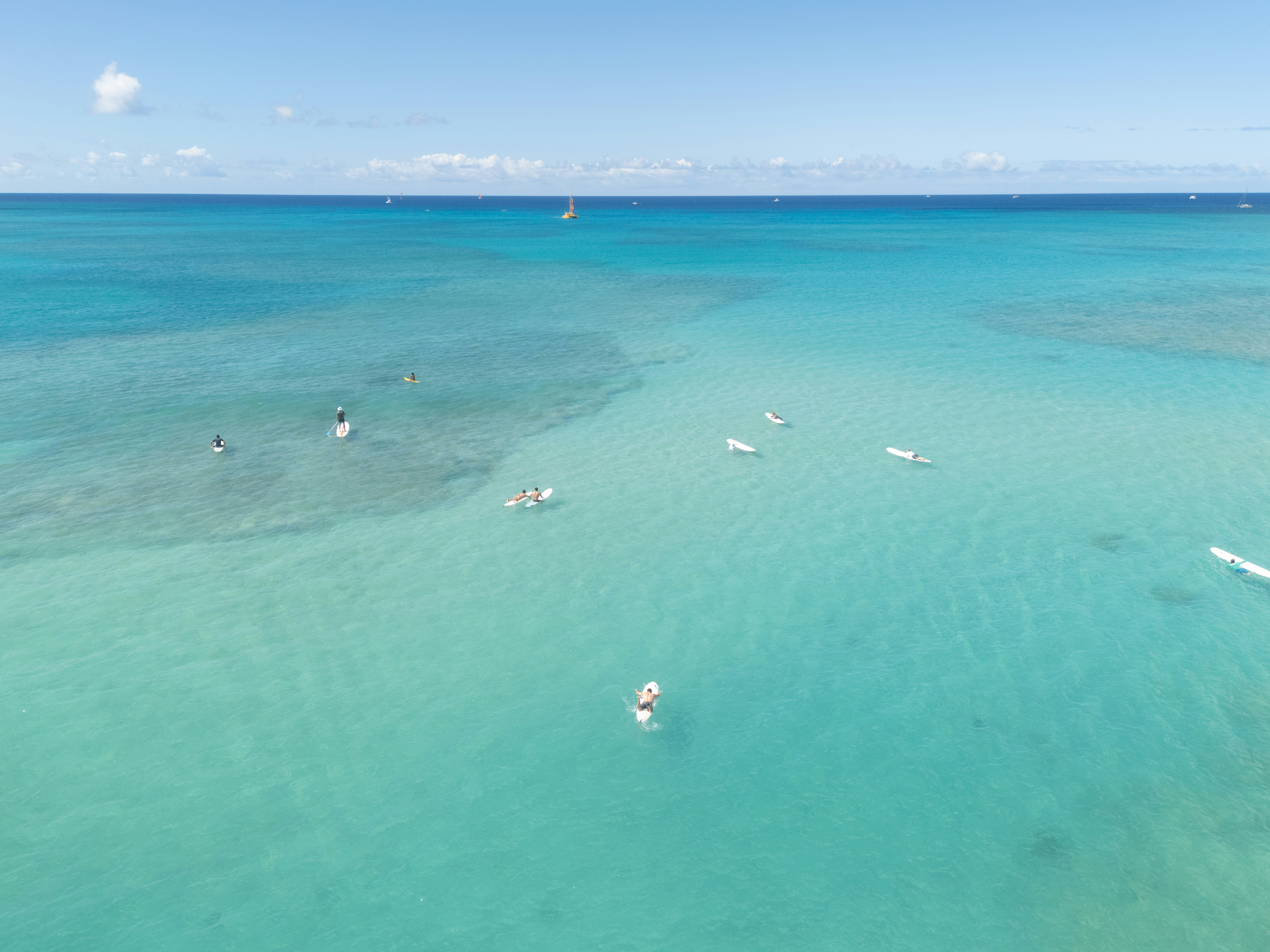 Aerial view of surfers and paddleboarders enjoying the calm, turquoise waters under a clear blue sky.