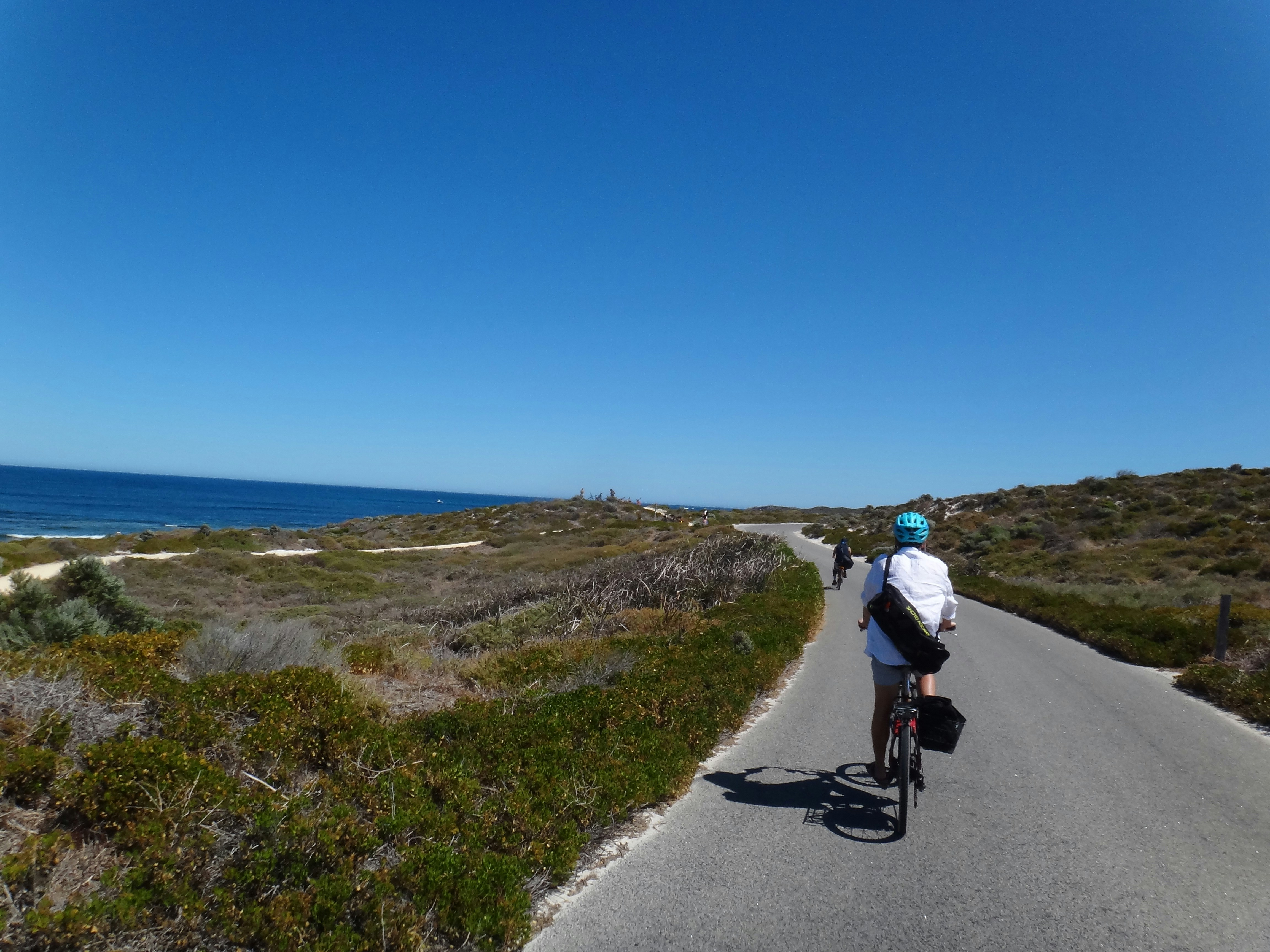 A cyclist rides along a sunlit coastal road with the sea on the left and scrubby dunes on the right.