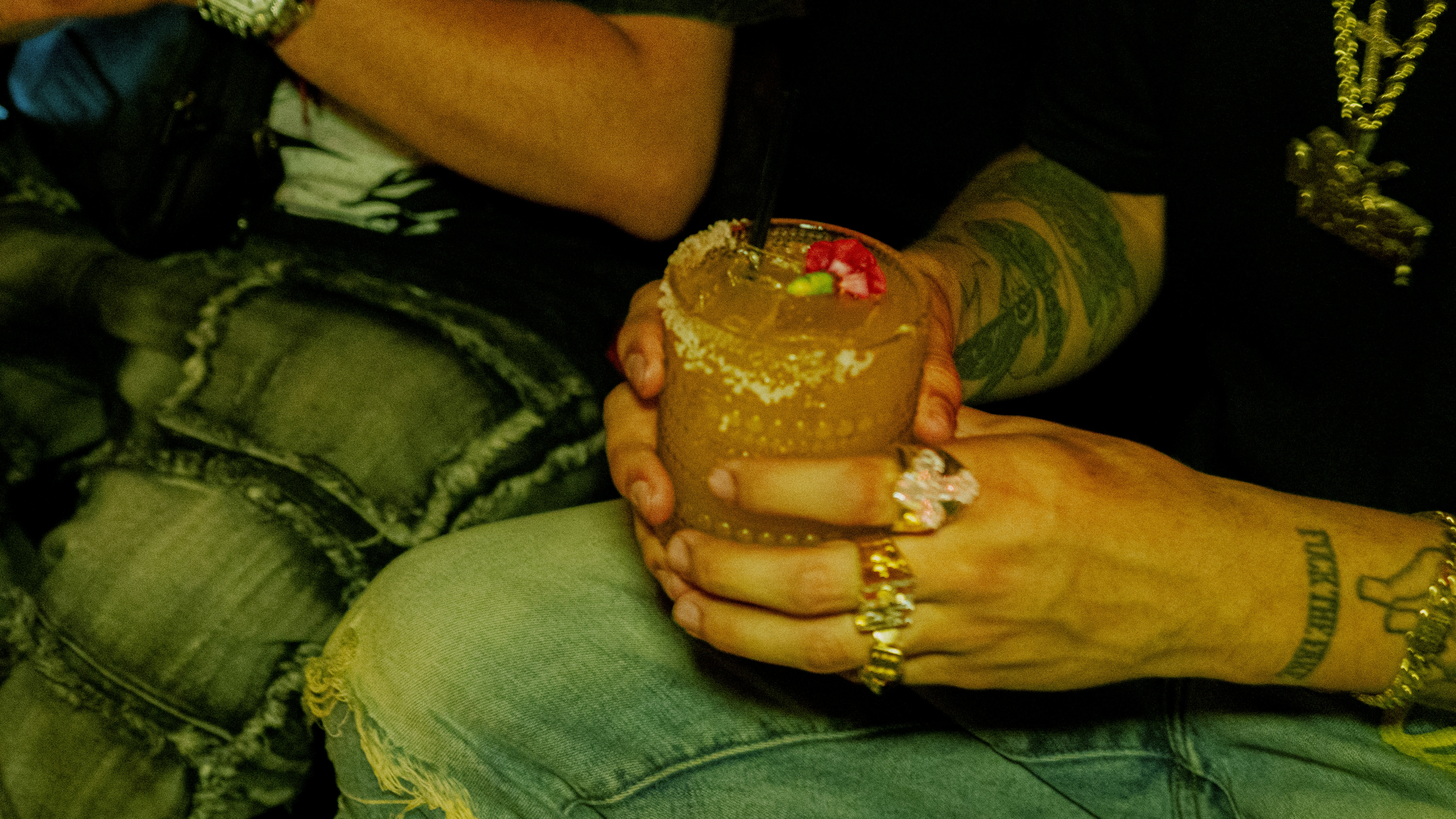 Gilded Sip: Tattooed Hands Grip a Citrusy Cocktail in Dim Light