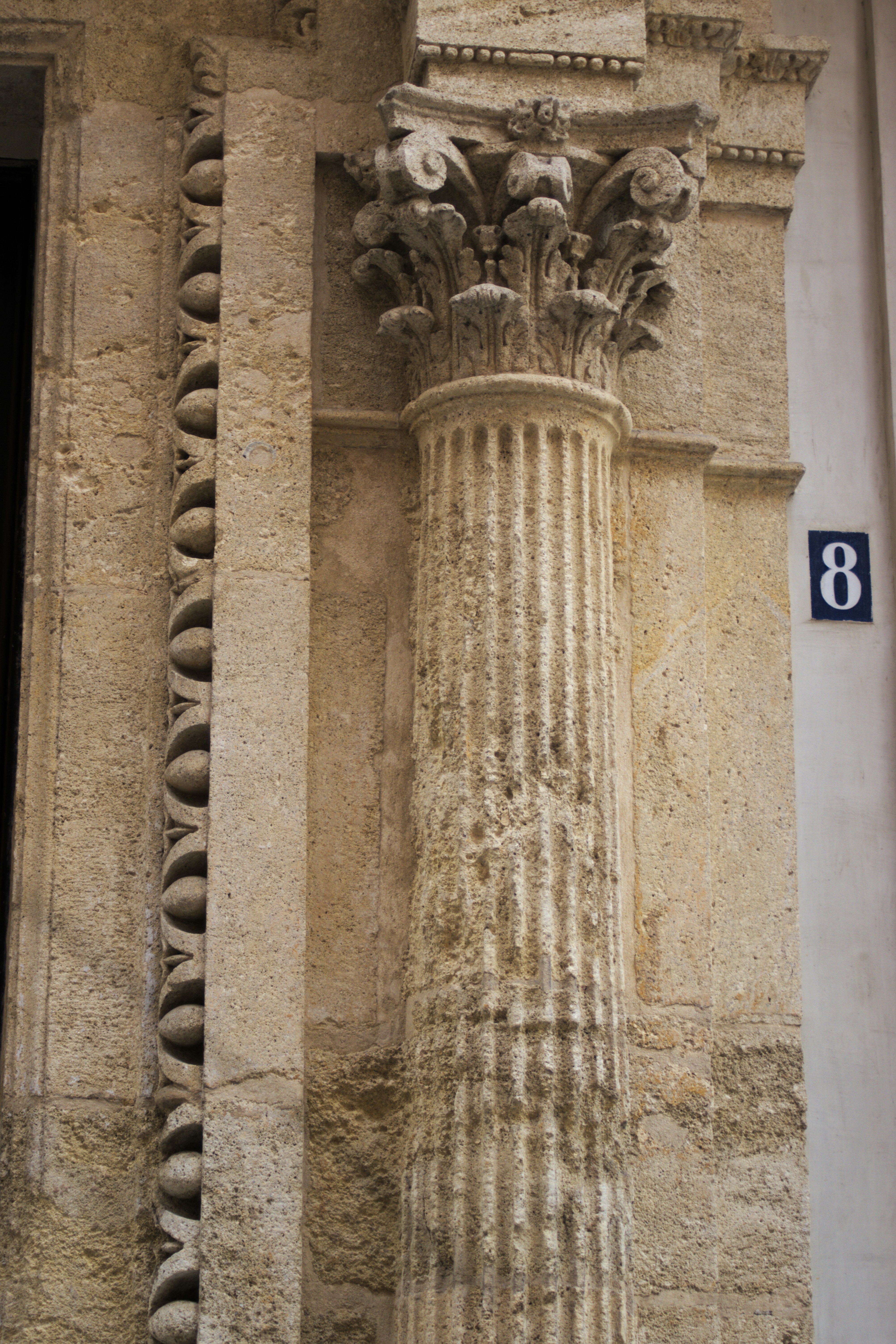 Silent Corinthian: Weathered Capital Framed by Ornate Stonework and a Crisp Blue Eight