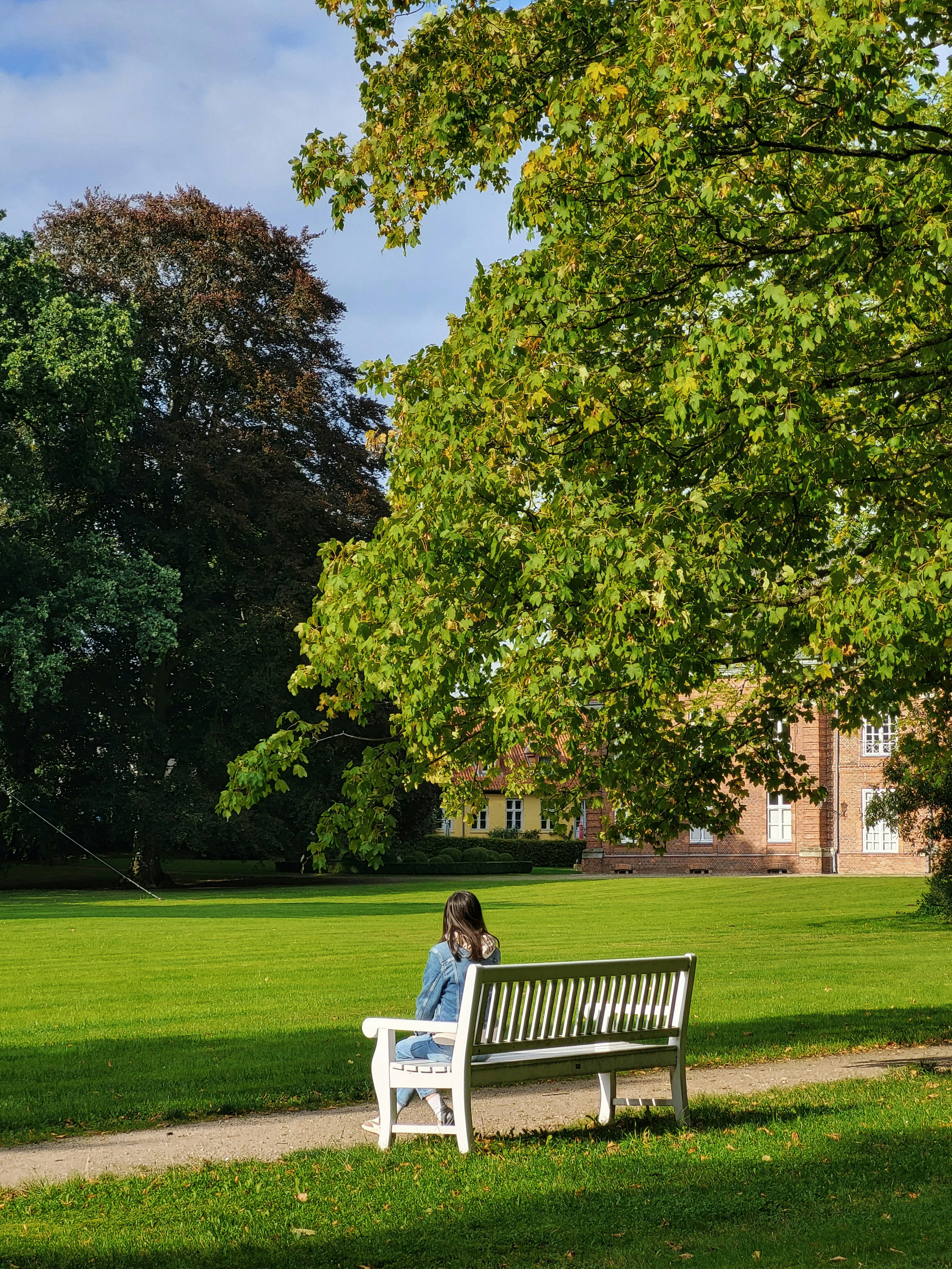 Person in a denim jacket sits on a white bench amidst green park with autumn trees and historic buildings.