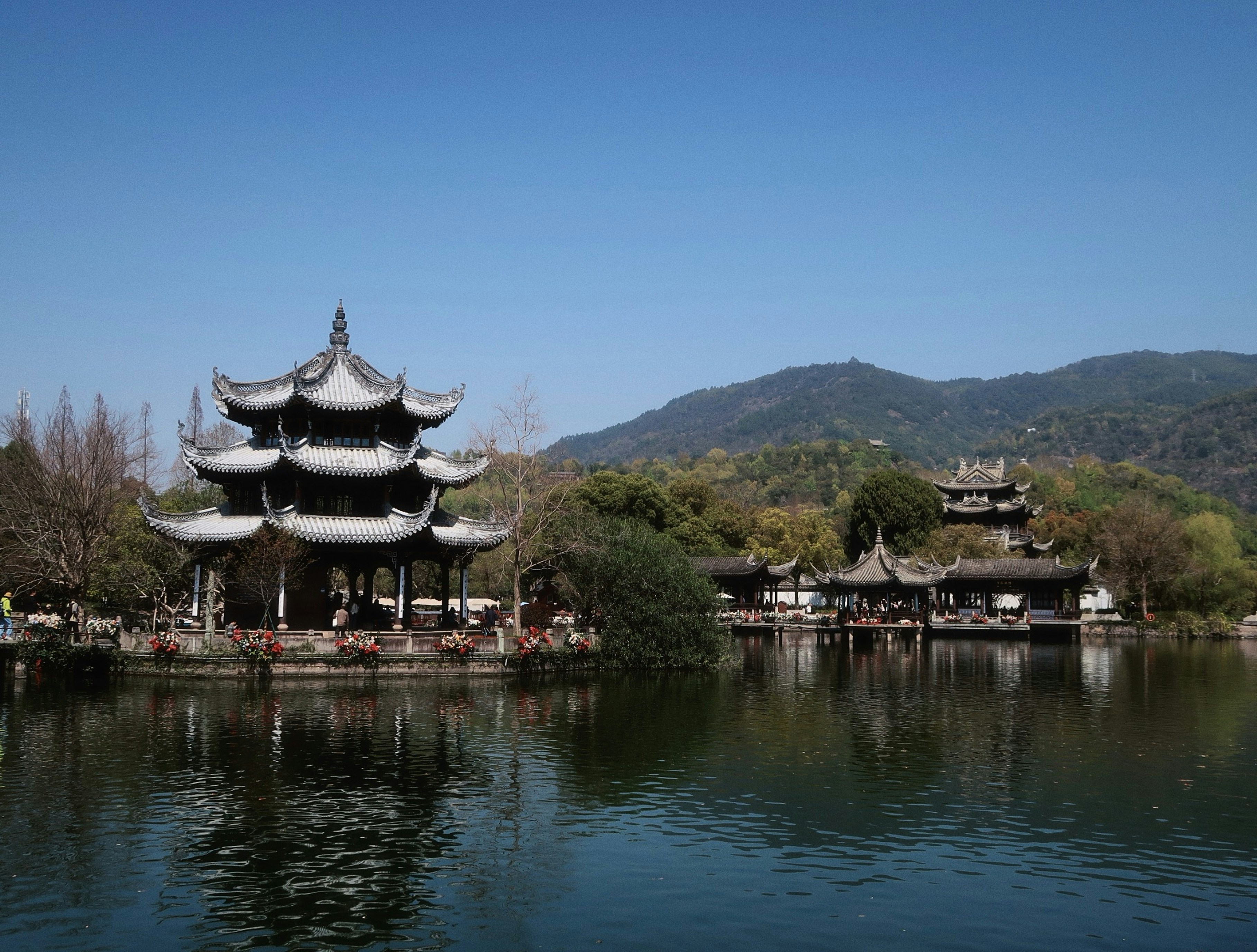 Traditional Chinese pavilions mirrored in a tranquil lake with distant mountains under a clear blue sky.