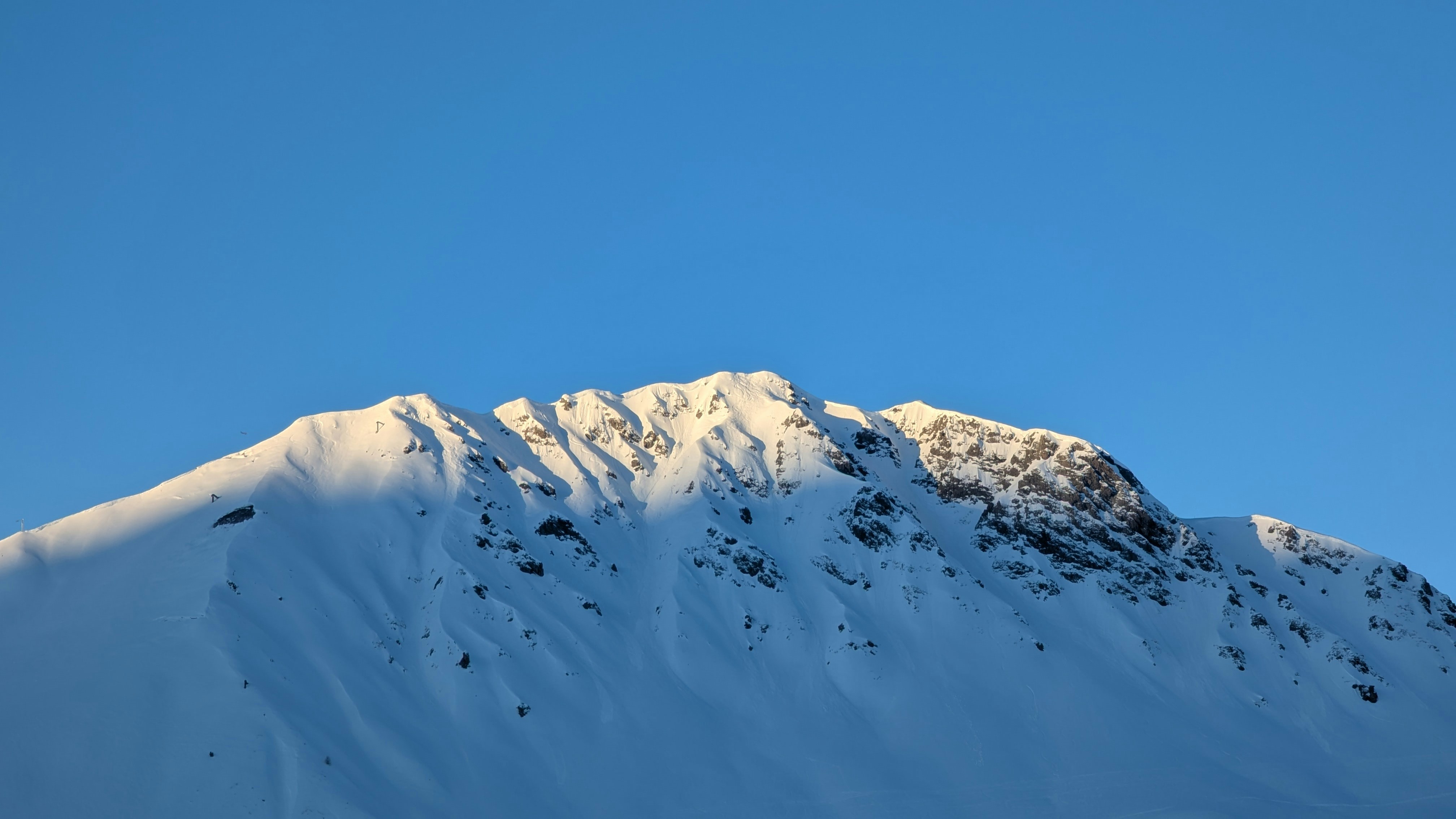 Snow-covered mountain peaks illuminated by sunlight under a cloudless blue sky.