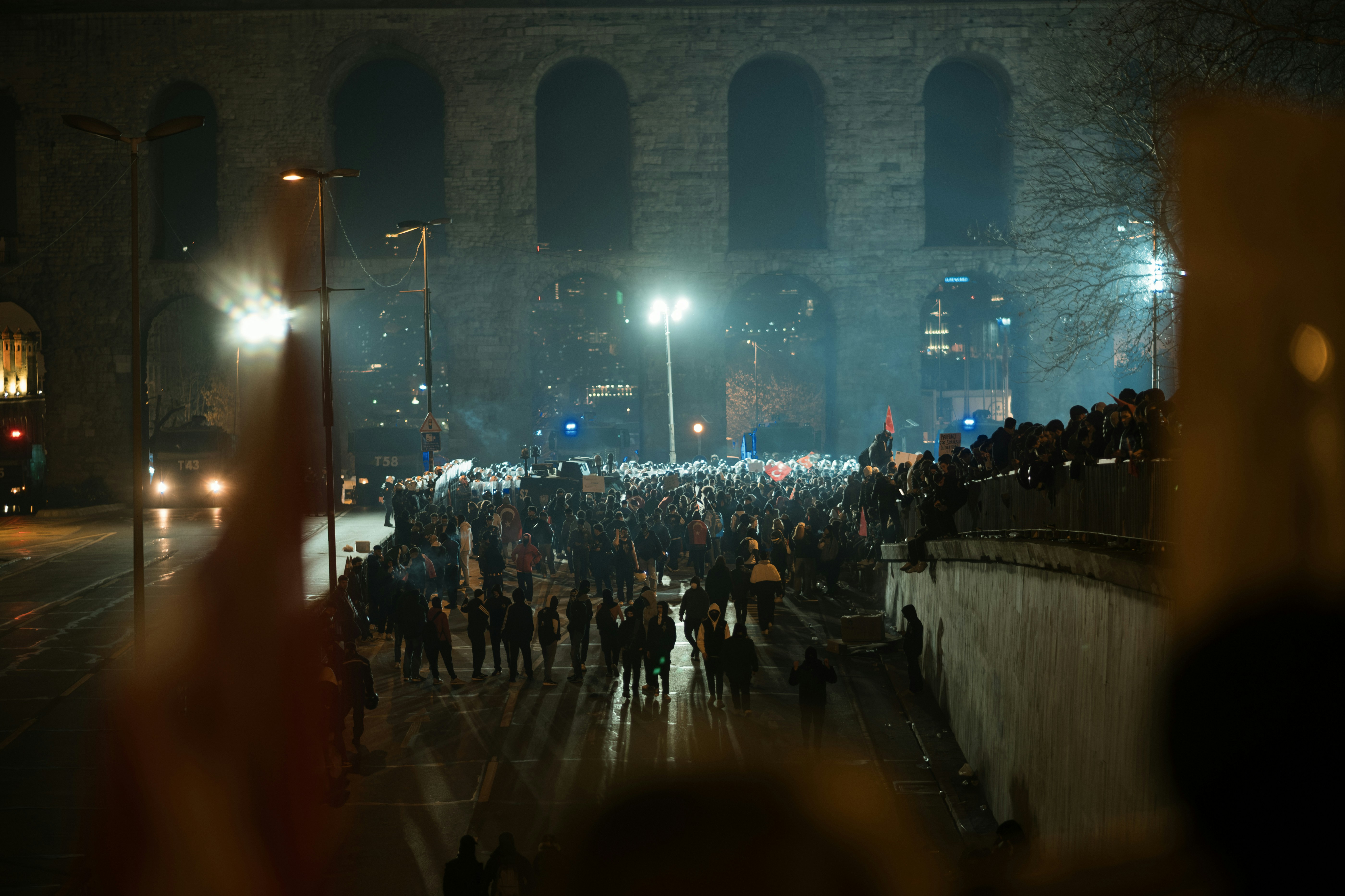 Crowd gathers near illuminated stone arches at night, with soft lighting and shadows creating a dramatic scene.