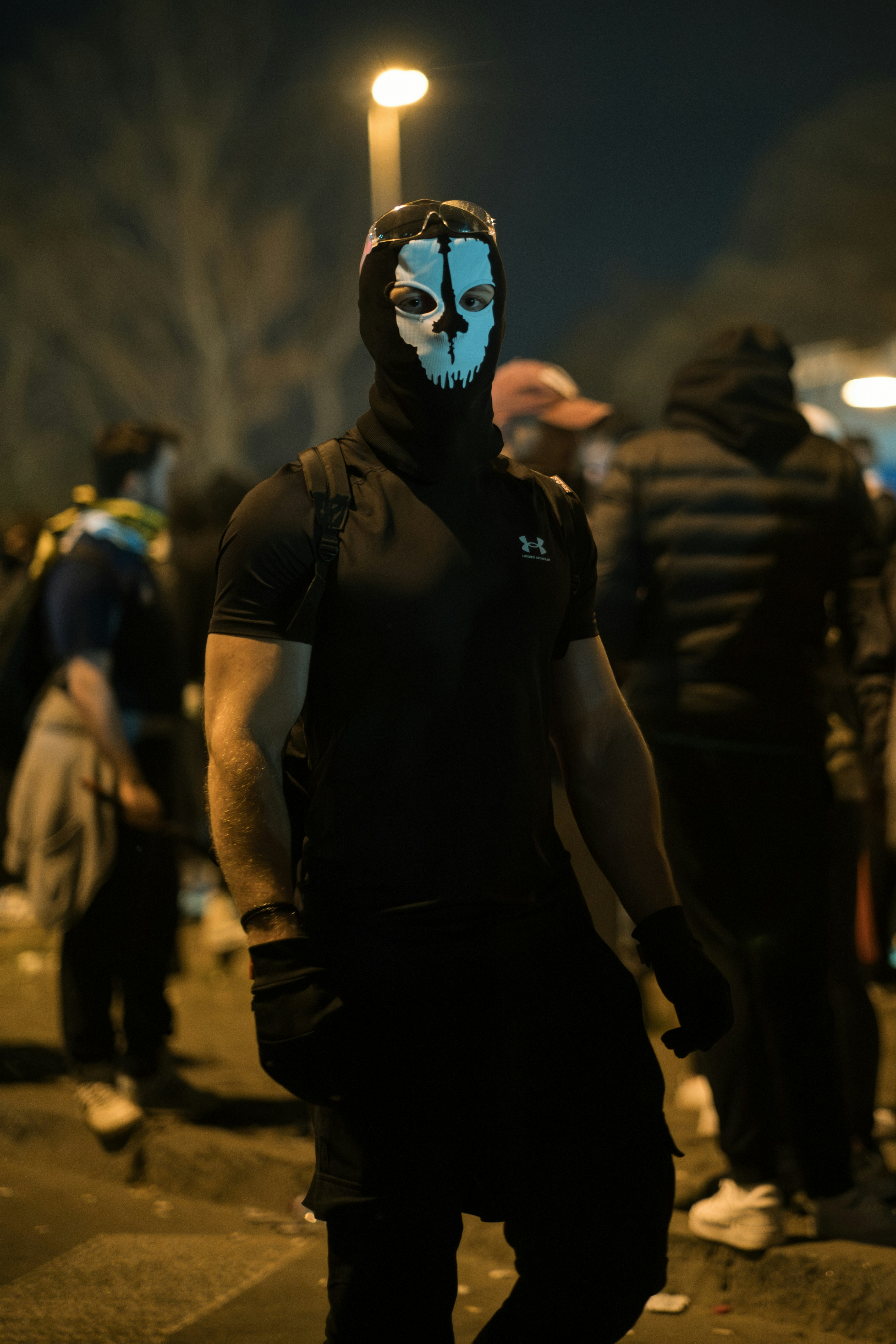 A figure in a dark mask stands resolutely amidst a crowd, embodying a sense of mystery and intent. The urban night setting adds to the atmosphere.