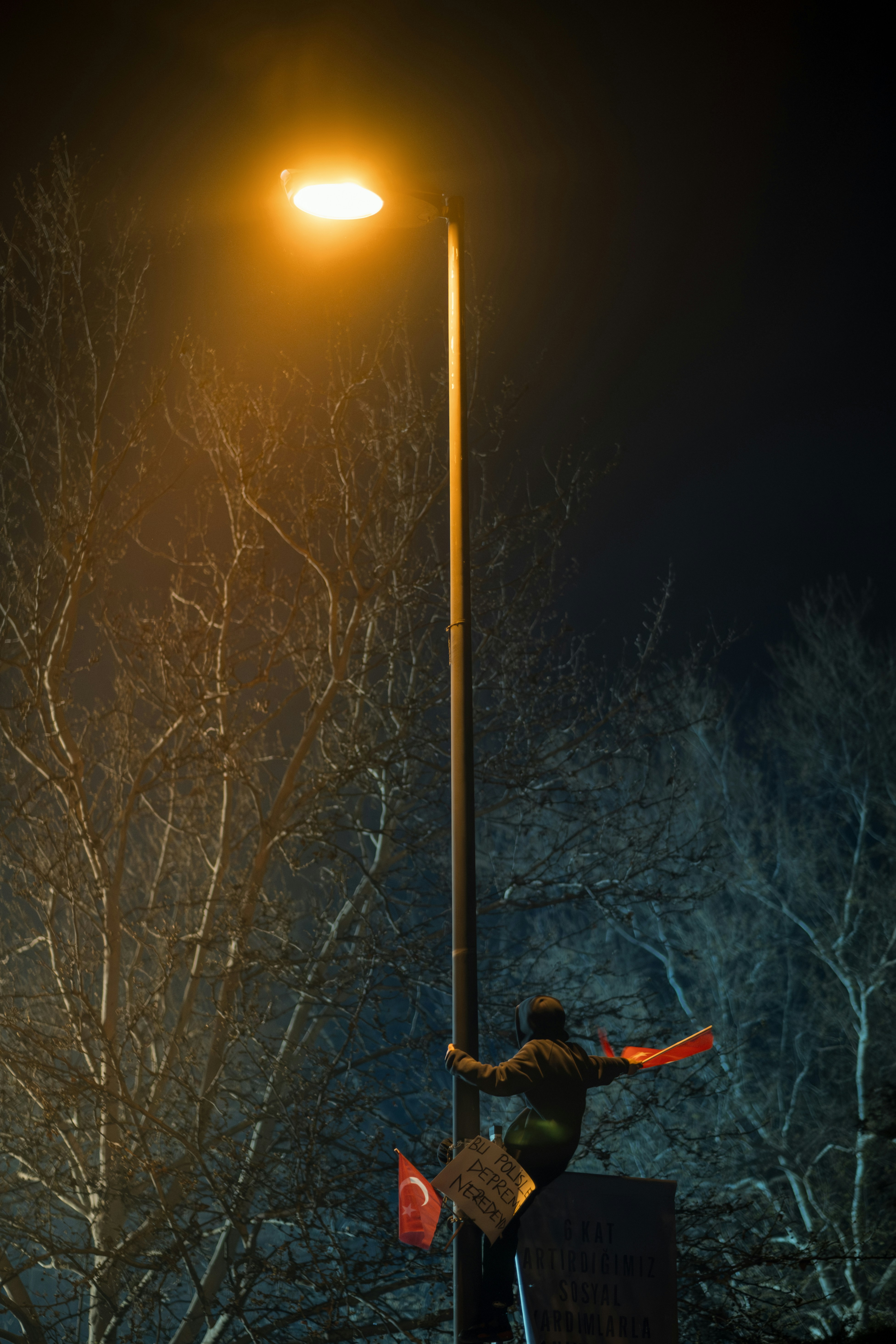 Lamppost Climber at Night: Flags, Shadows, and Amber Glow