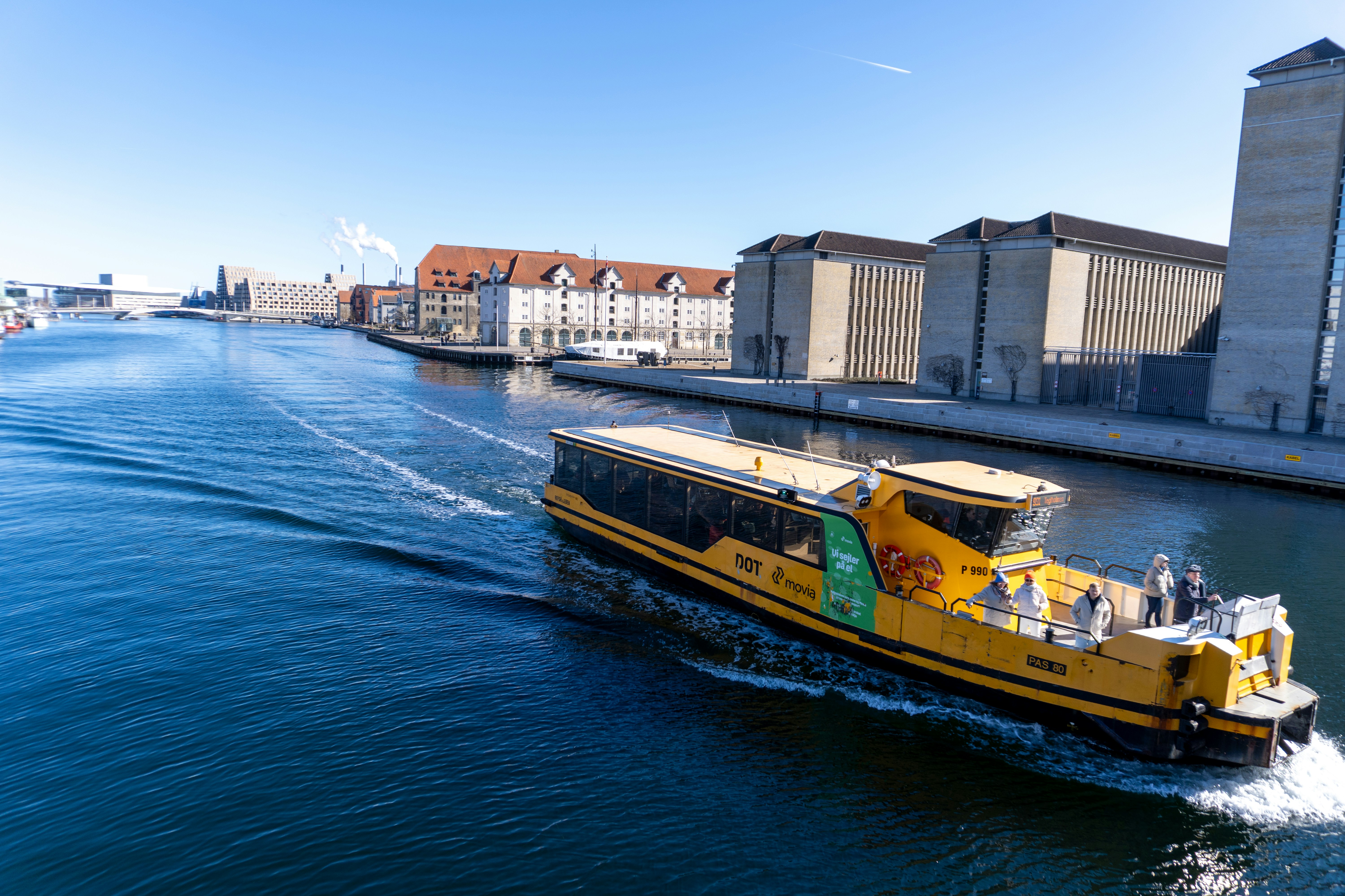 Yellow water taxi gliding through a sunlit canal with industrial buildings lining the shore.