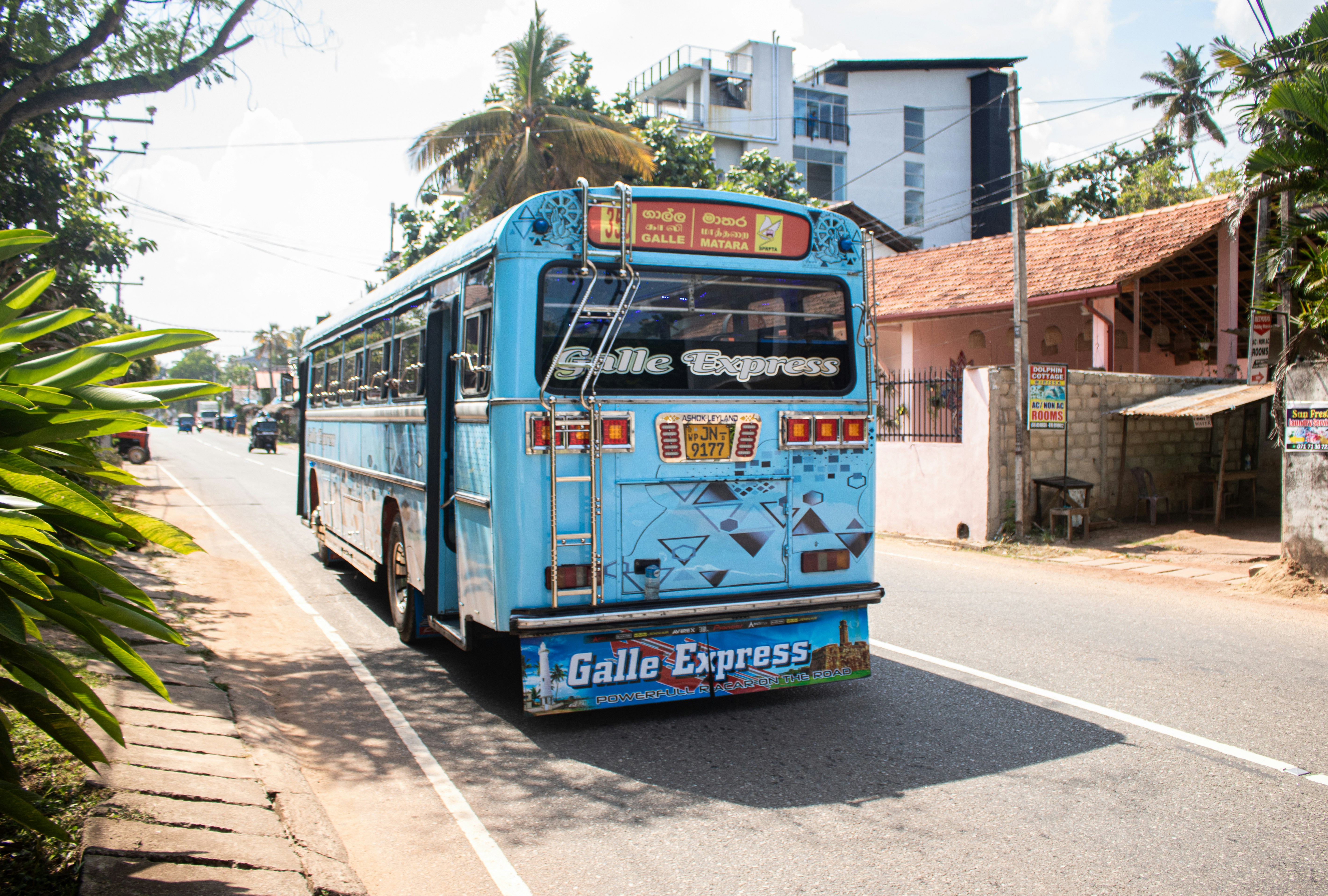 Light blue bus with 'Galle Express' branding travels down a sunlit street lined with palm trees and buildings.