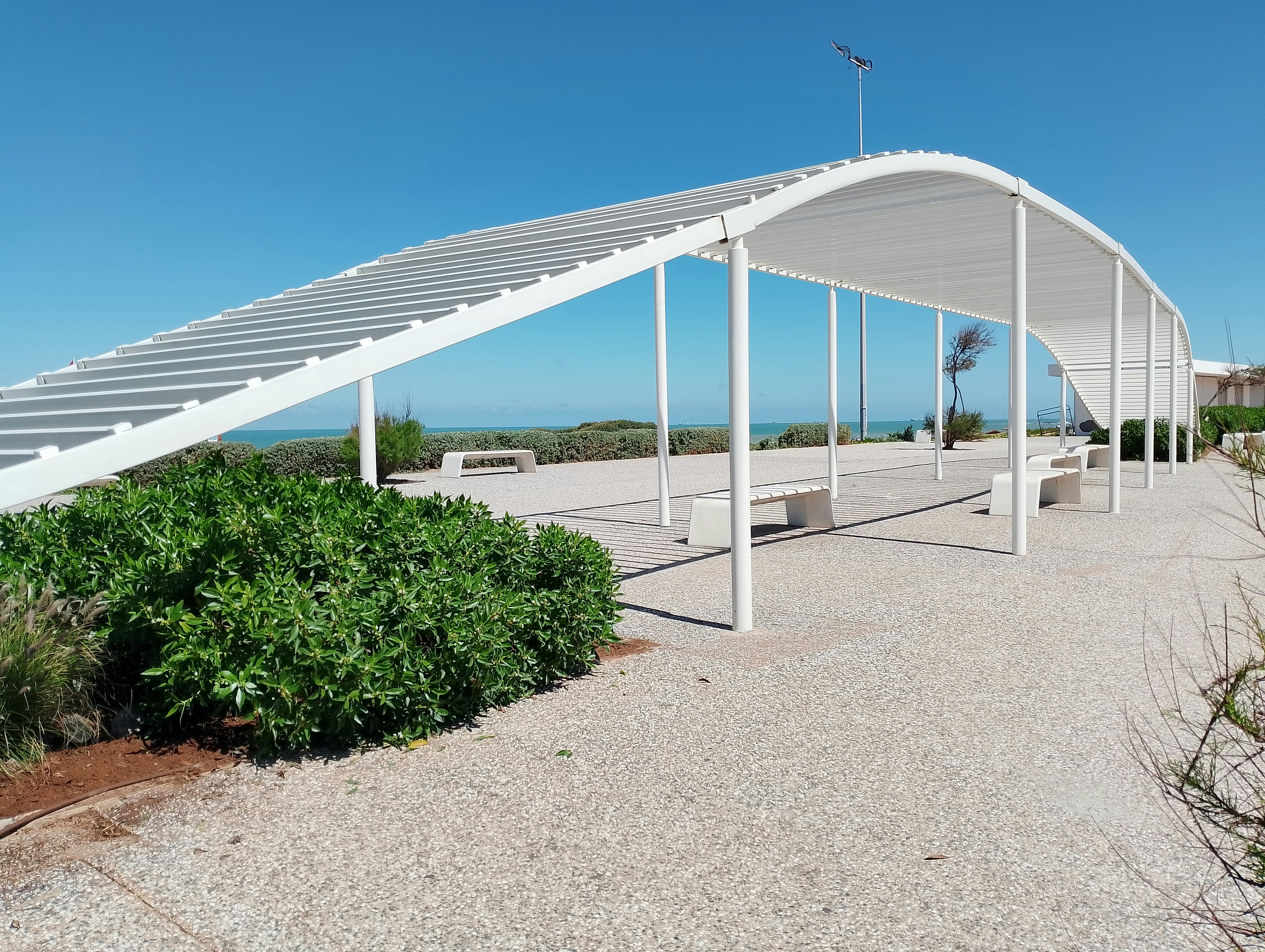 Modern white canopy structure with benches on a sunny coastal promenade.