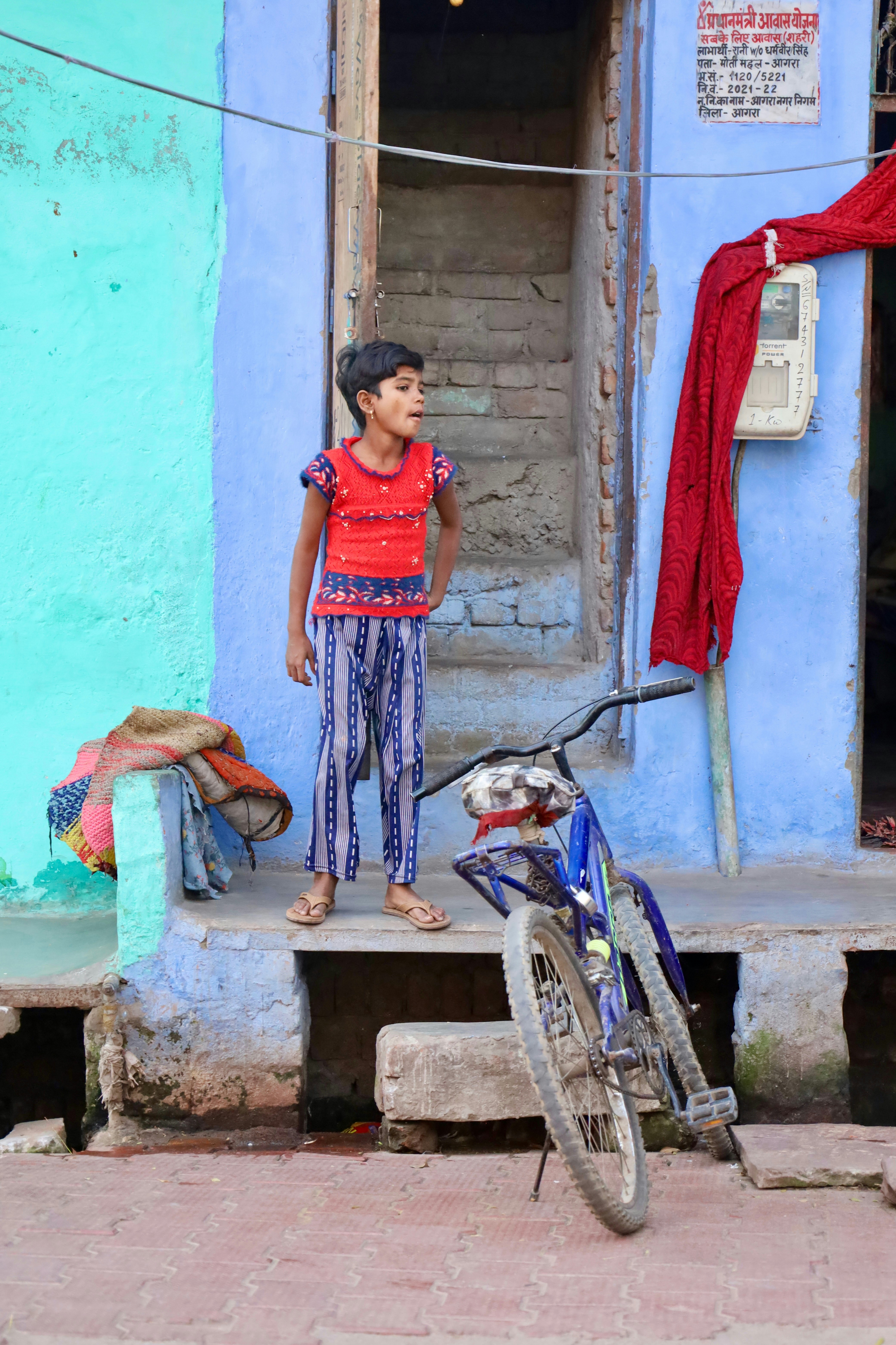 Child in colorful attire stands beside a tilted bicycle against a vividly painted wall.