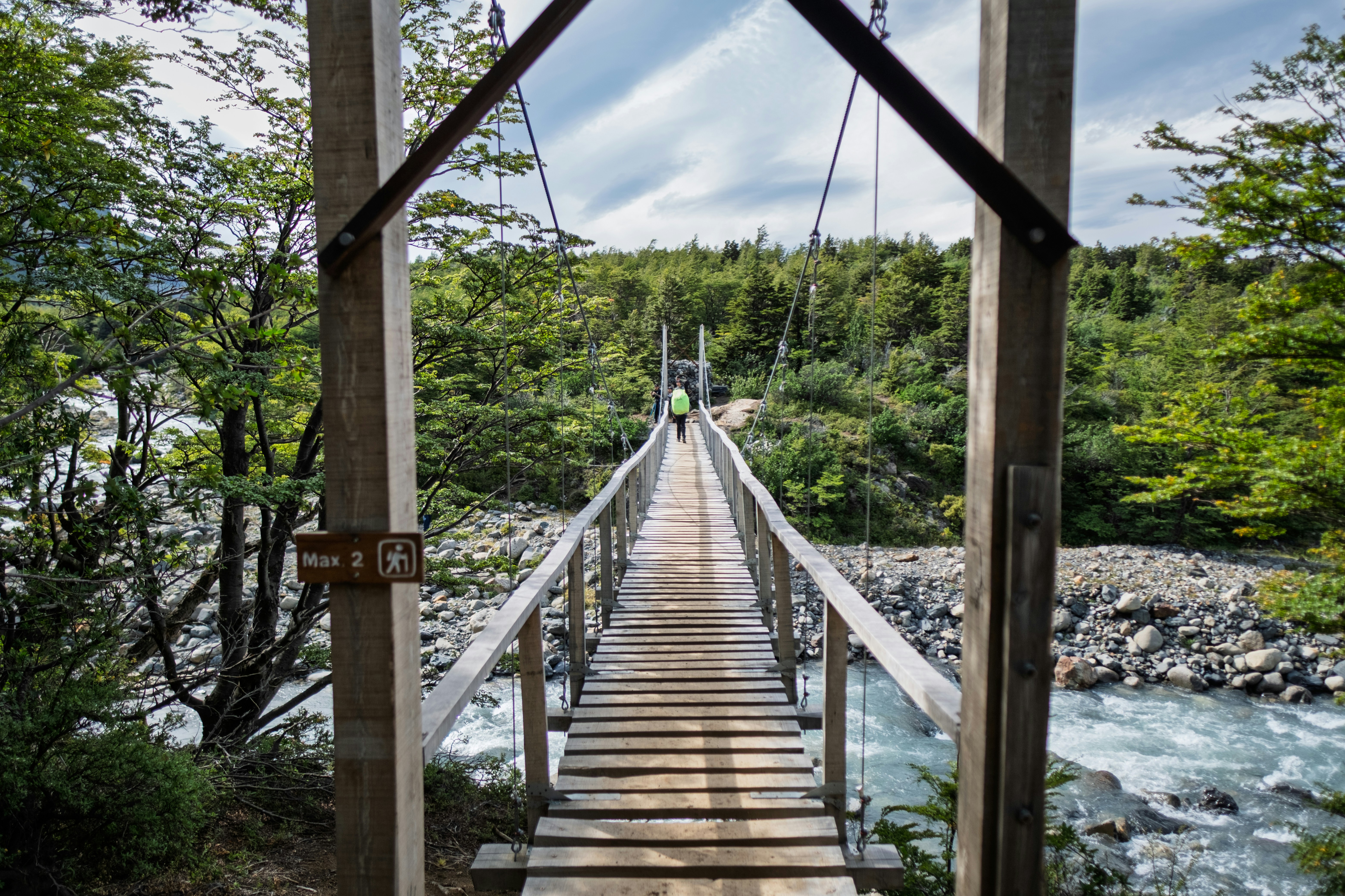 Wooden suspension bridge spans a flowing river, framed by lush green foliage under a dynamic sky.
