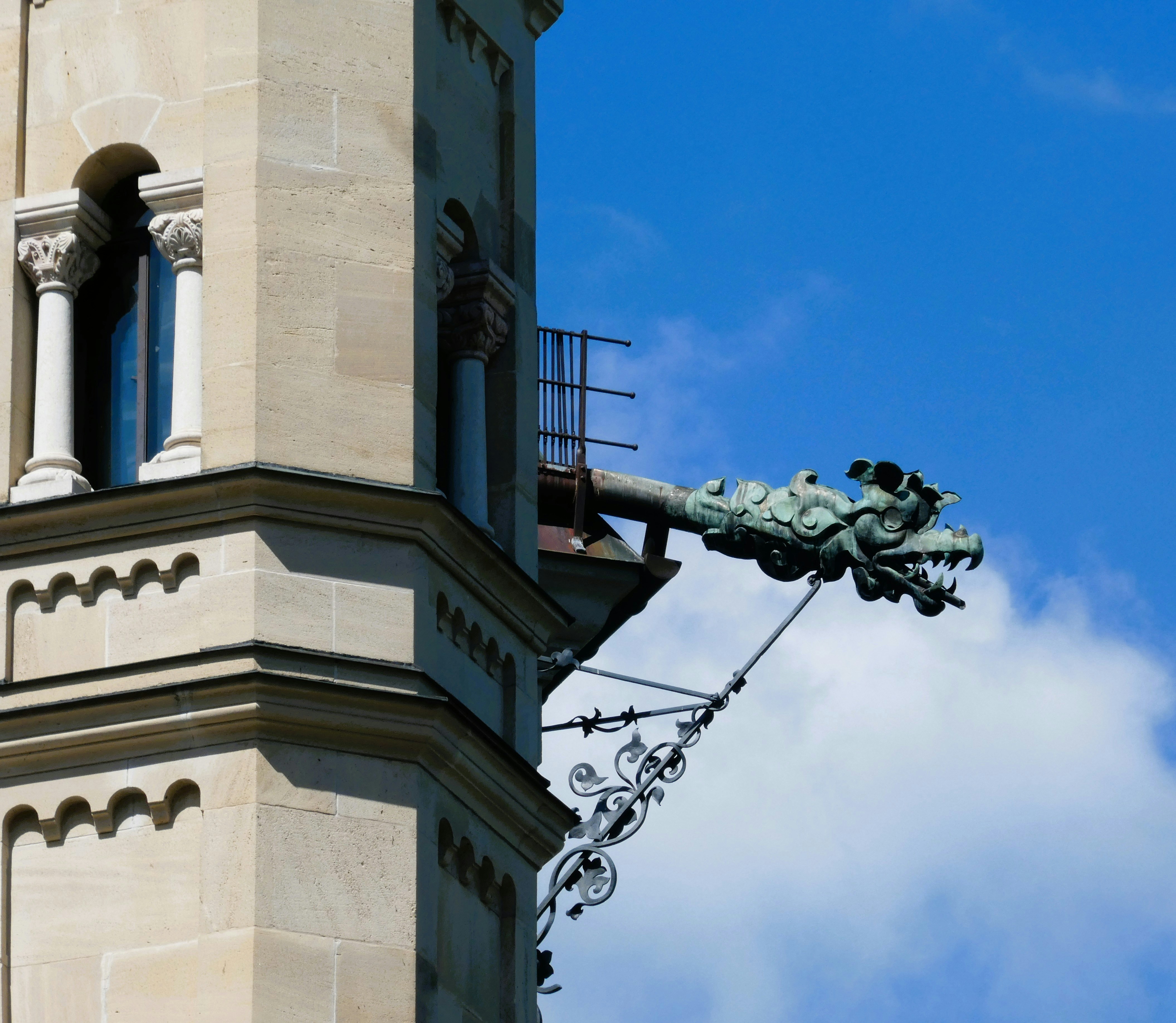 Architectural detail of a stone spire featuring an ornate dragon gargoyle extending outward. The sculpture stands against a bright blue sky, highlighting craftsmanship and weathered stone.