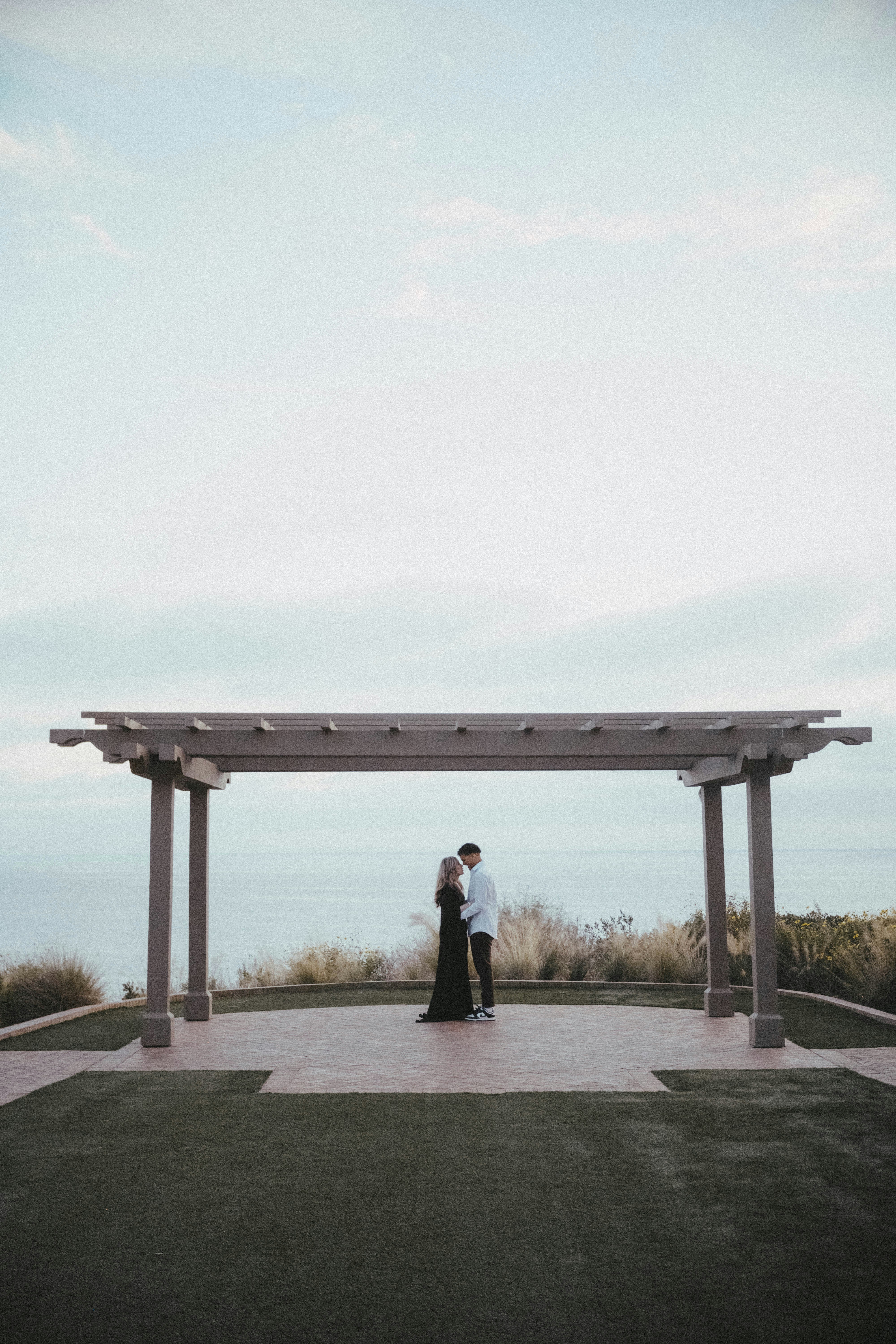 Couple standing under a pergola on a terrace overlooking the sea with a pastel sky.
