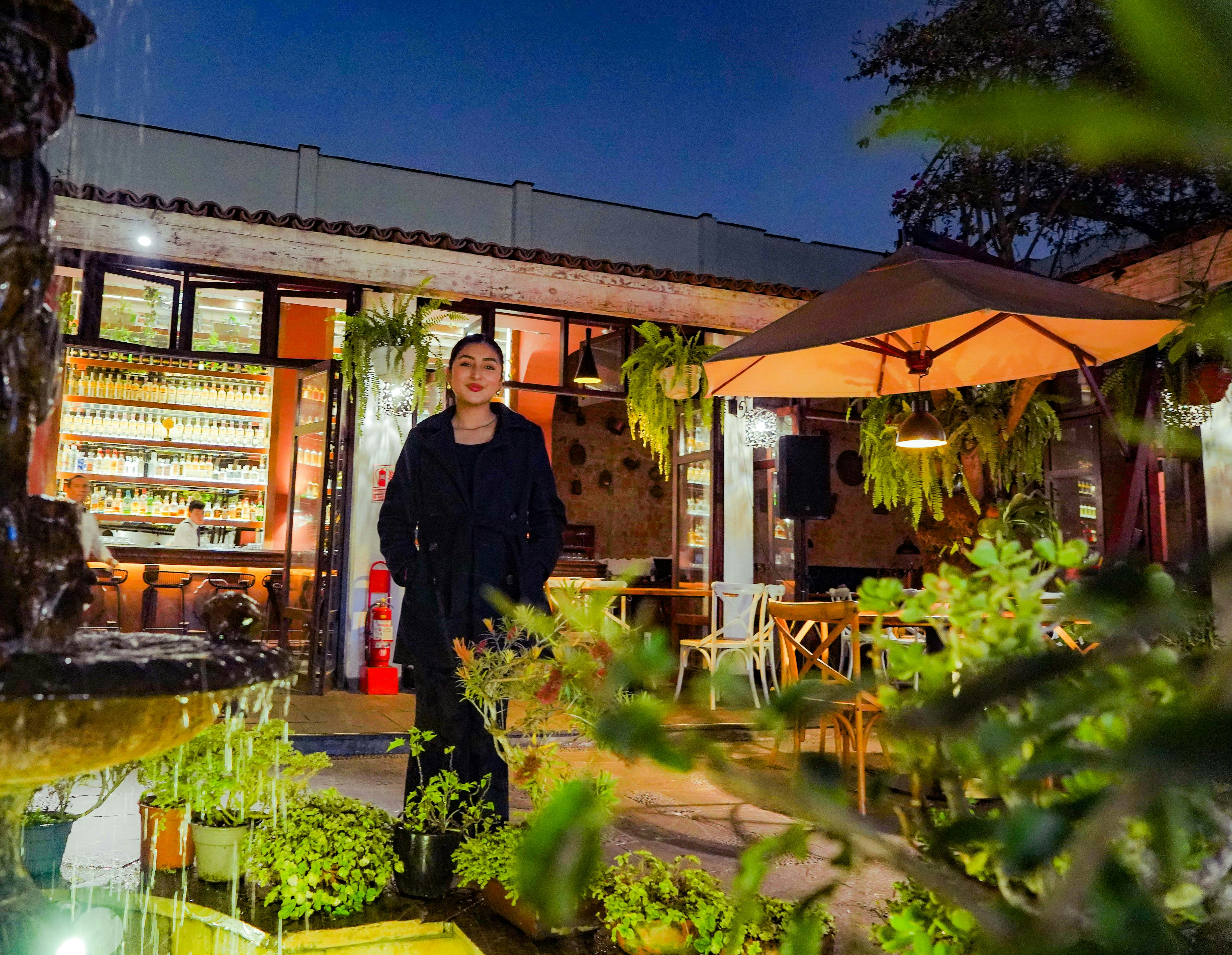Person standing in a cozy outdoor café surrounded by lush greenery and warm lighting at dusk.