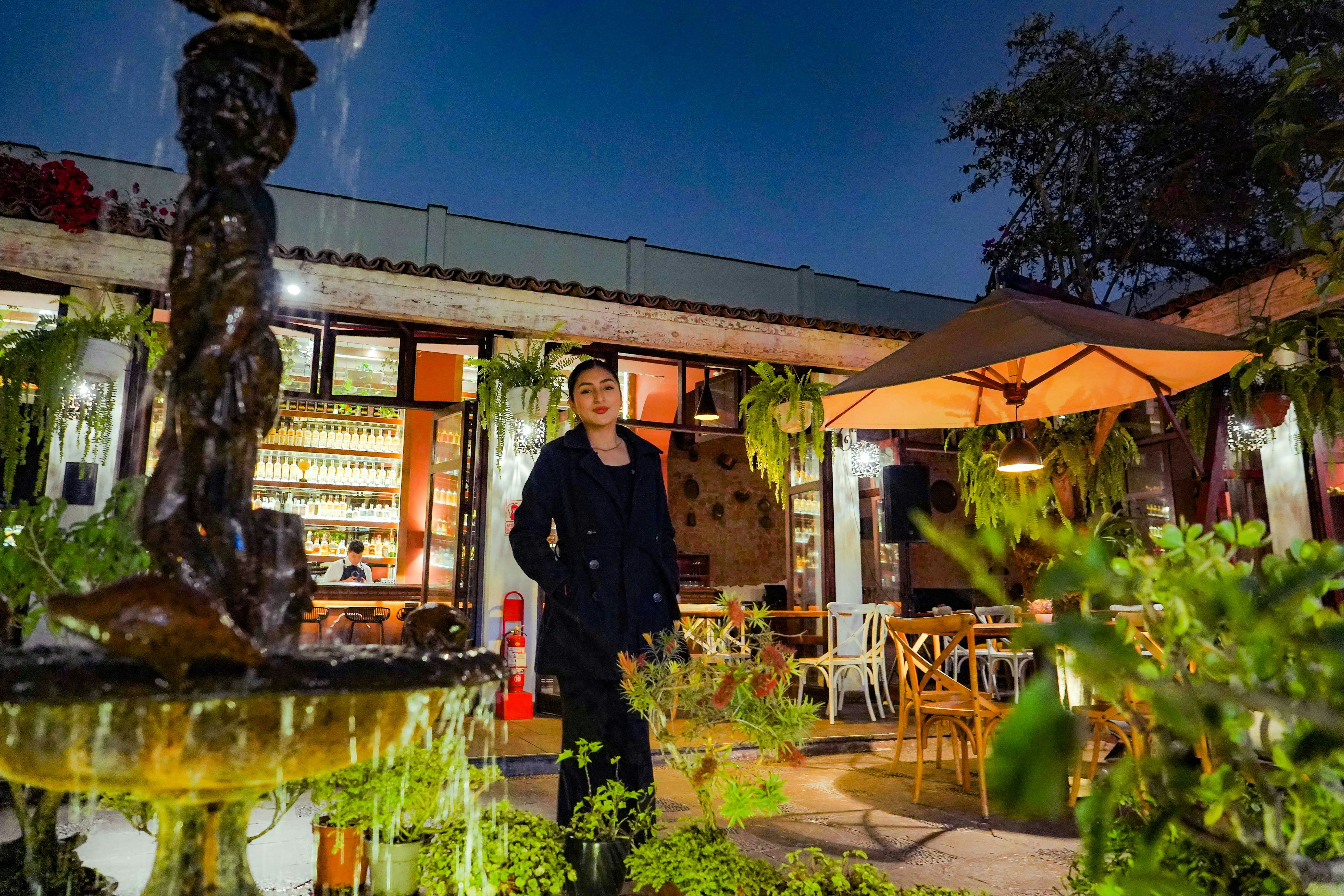 Man standing near a fountain in a lush outdoor café setting under a twilight sky.