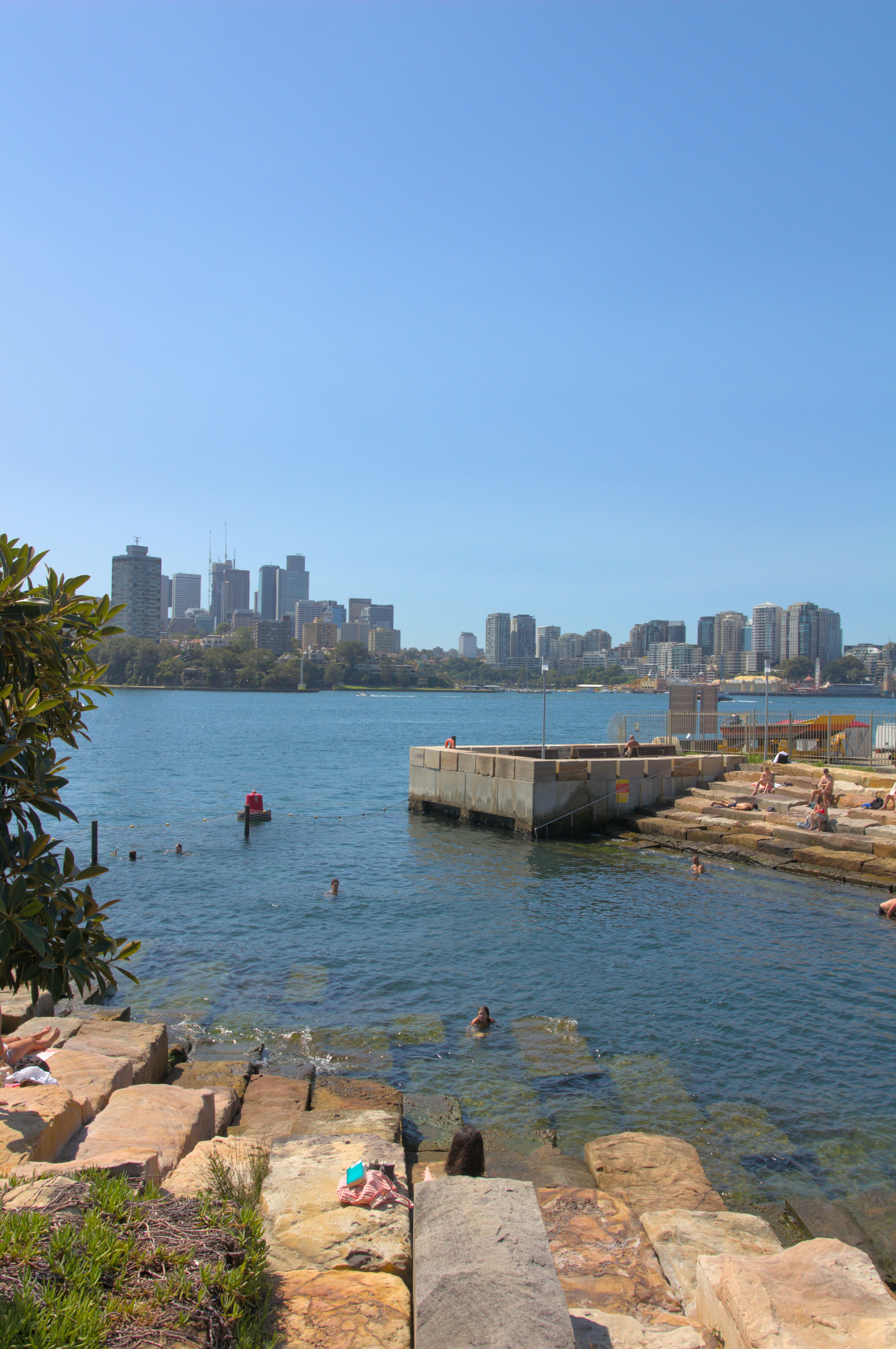 Sunlit urban skyline viewed from a rocky shoreline with swimmers enjoying the water.