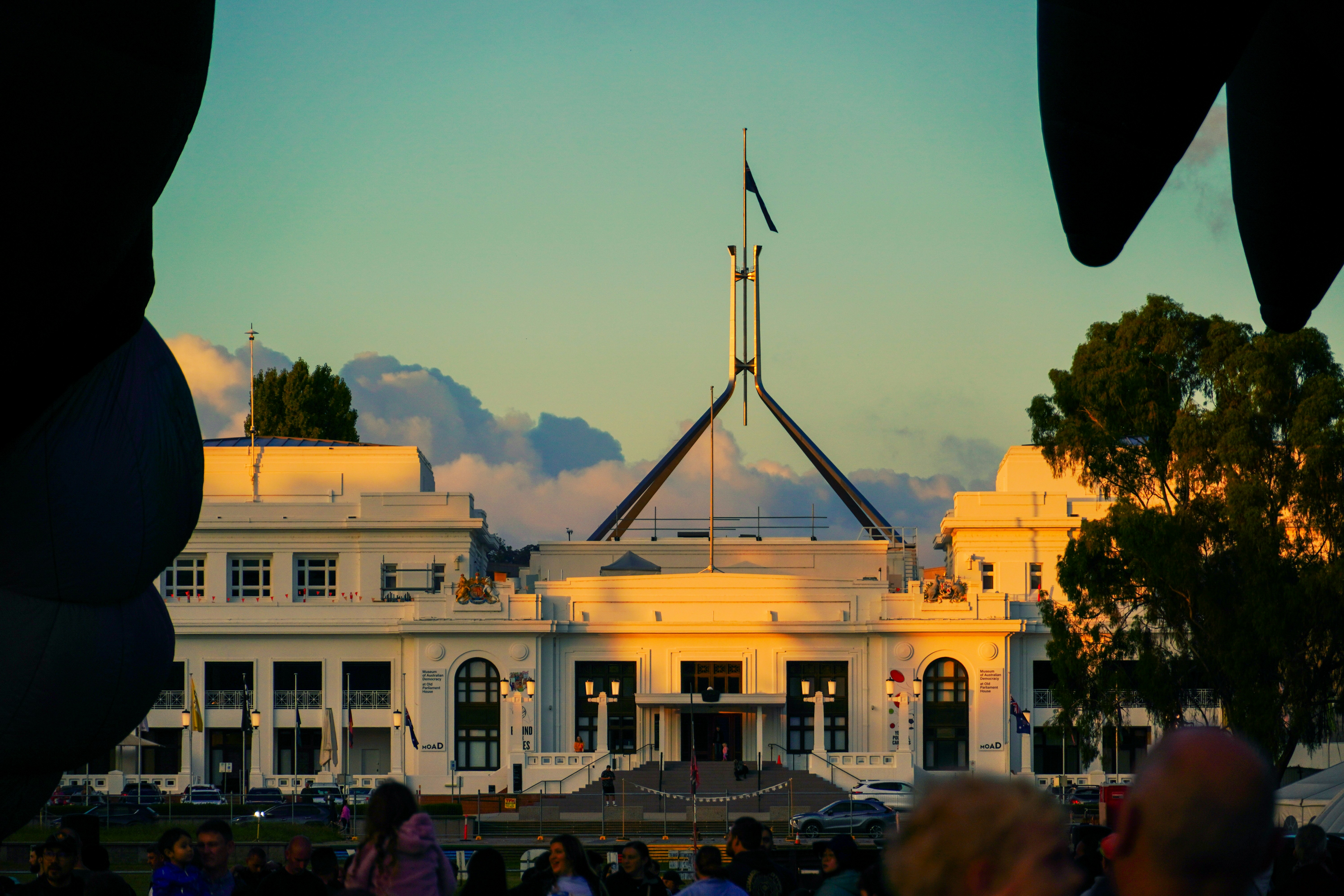 Parliament House of Australia illuminated by the golden light of the setting sun, with people gathered in front.