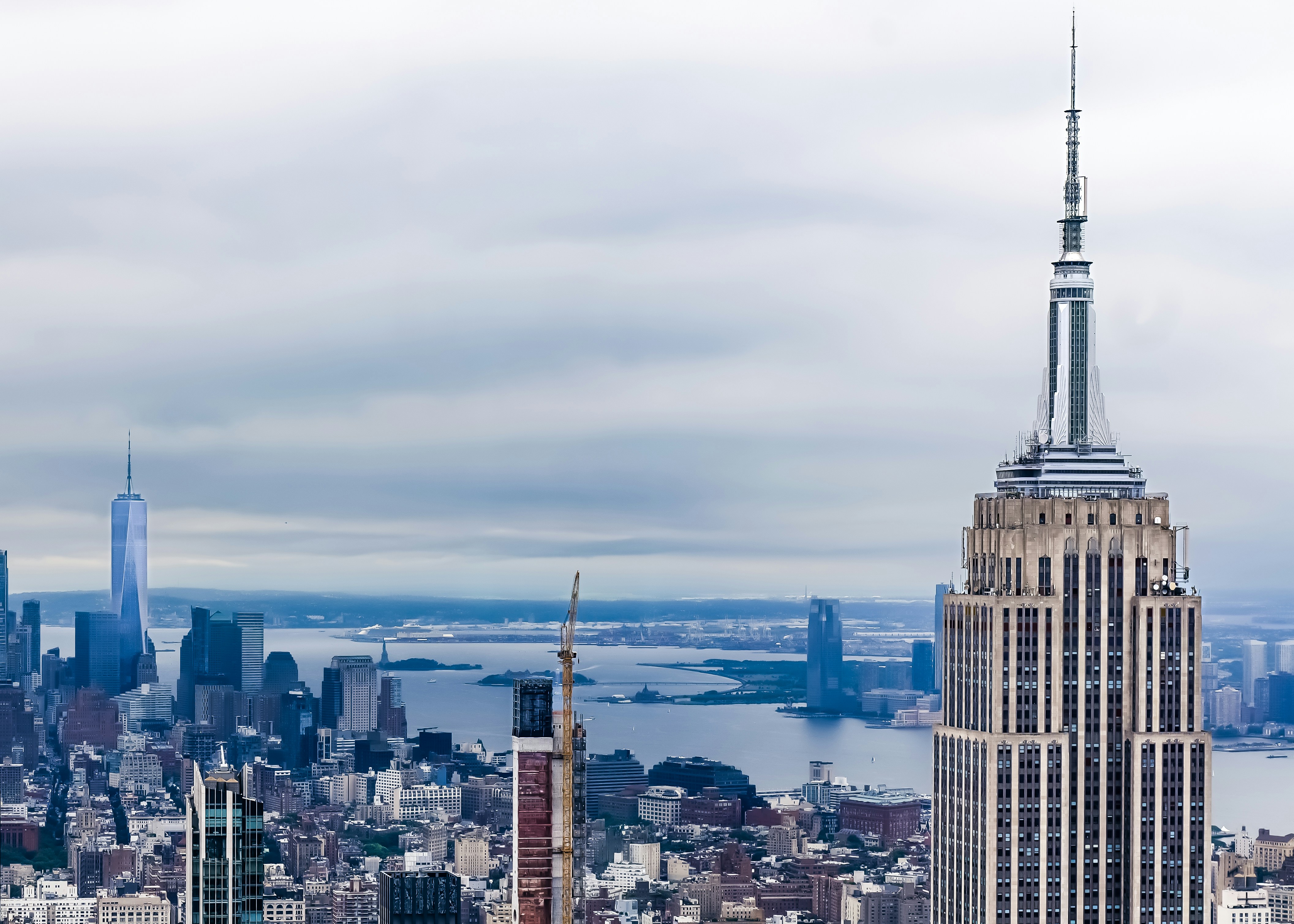 Empire State Building towering over New York City with a backdrop of the Hudson River and distant skyscrapers under a cloudy sky.