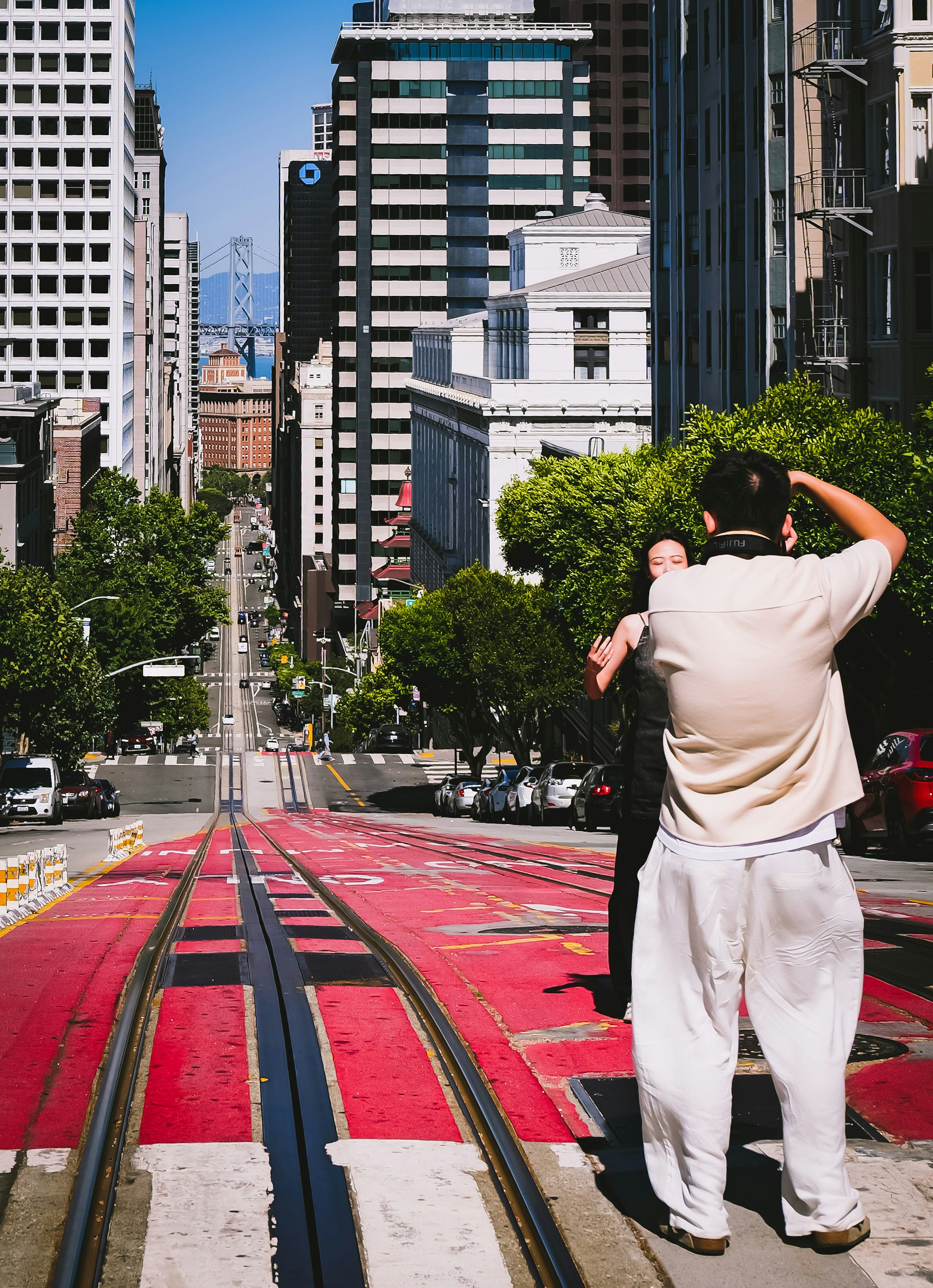 Photographer capturing bustling cityscape from a sloped street with vibrant tram tracks and towering buildings.