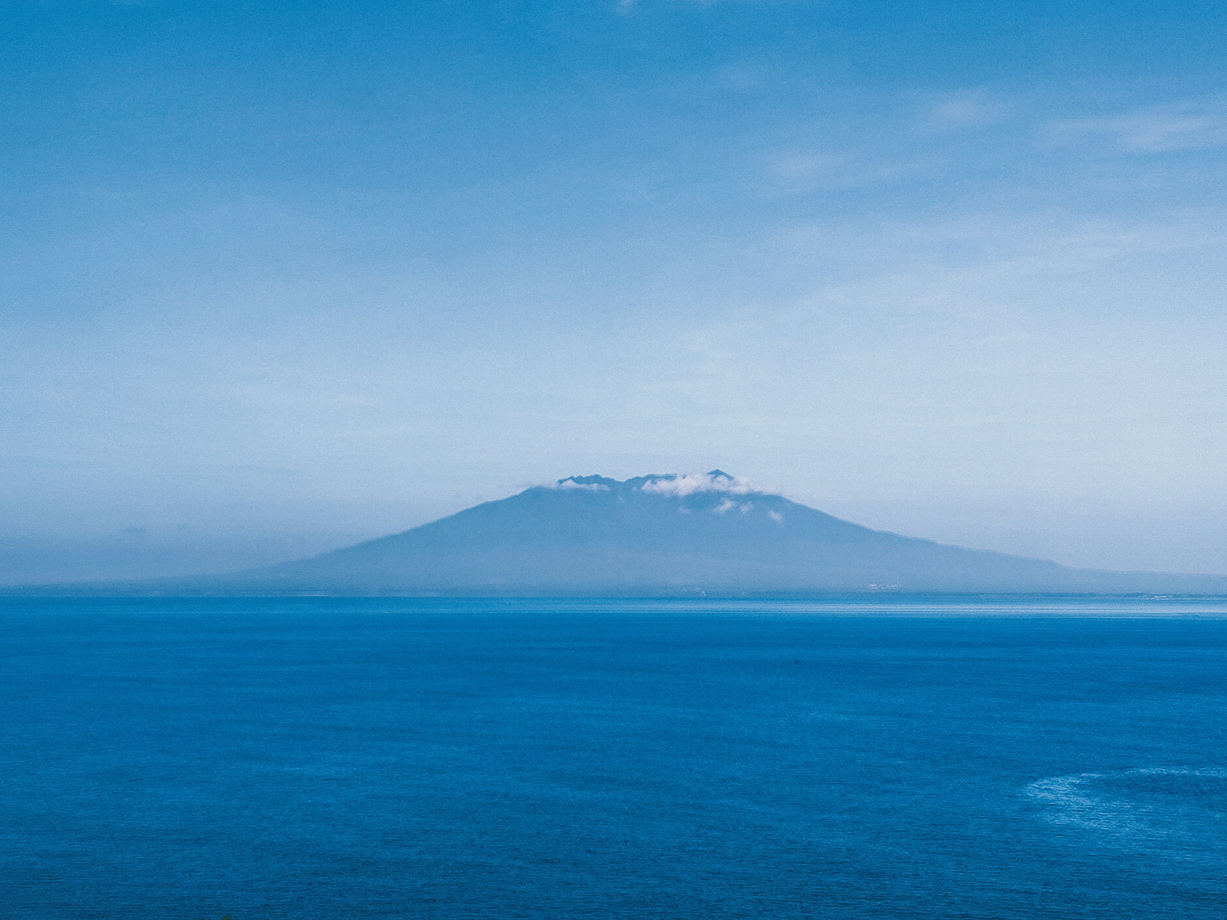 Misty mountain rising above a vast expanse of calm sea under a clear blue sky.