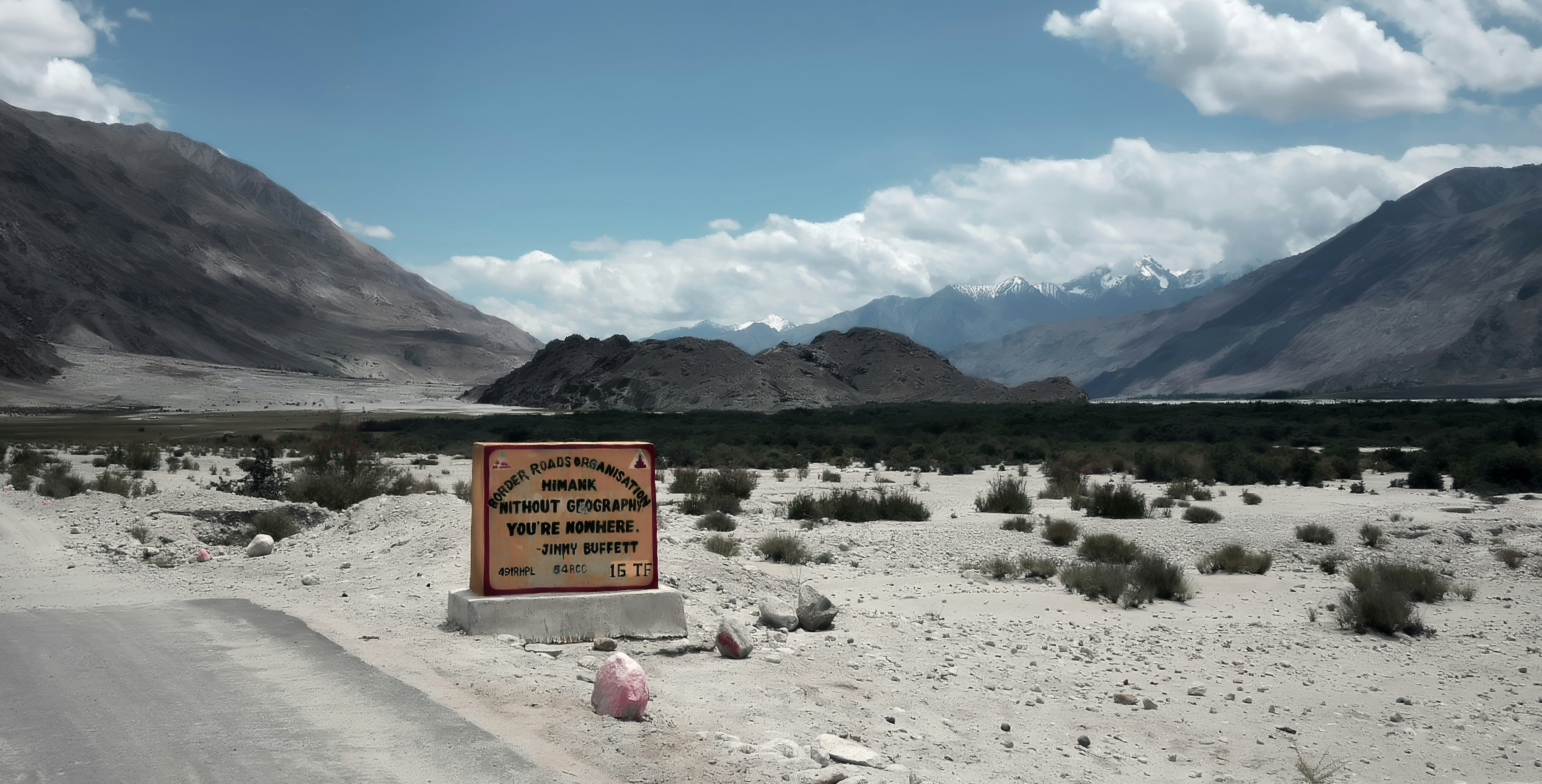 Weathered road sign in Nubra Valley's expansive, arid landscape with distant snow-capped peaks under a bright sky.