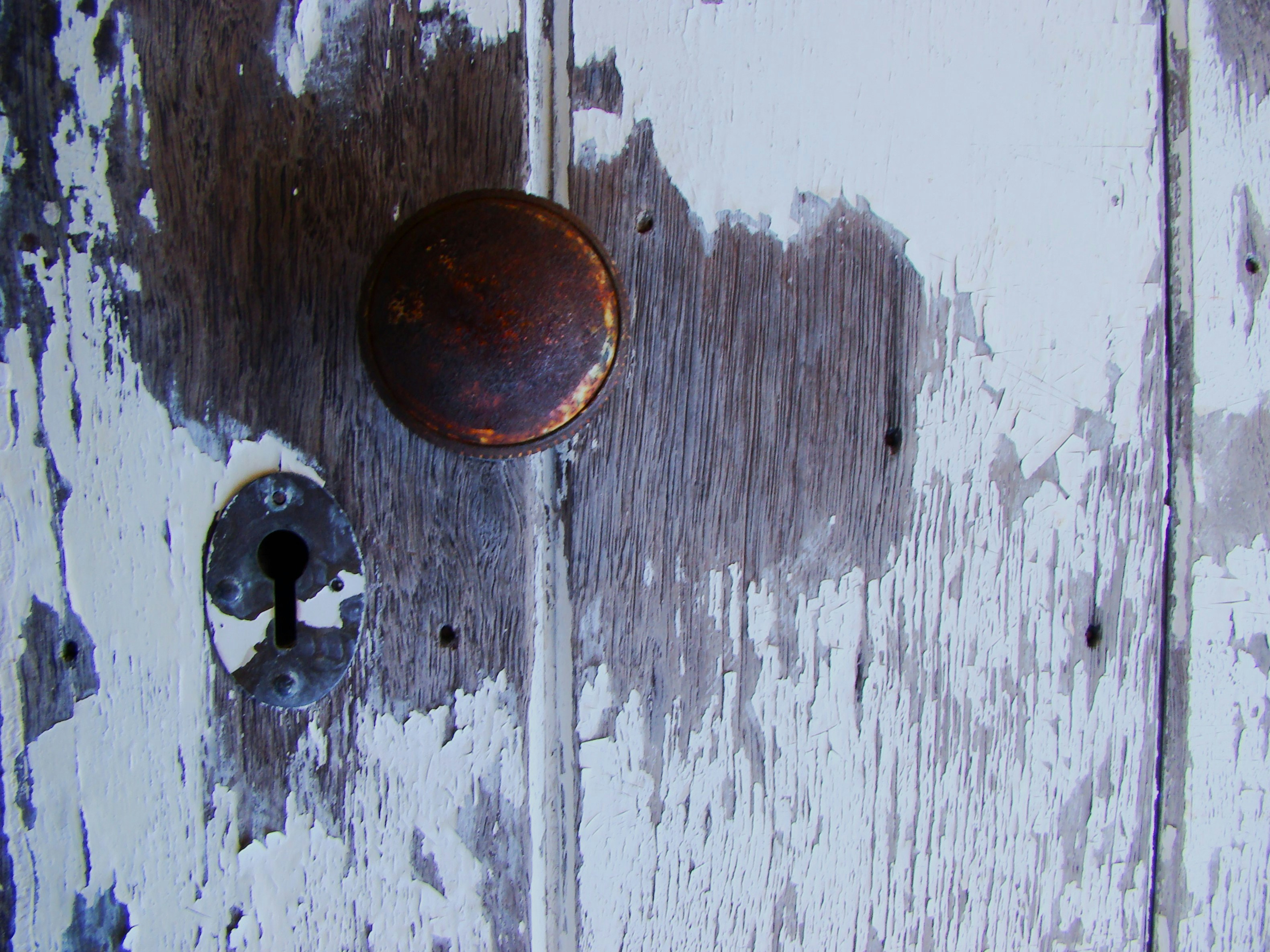 Close-up of a weathered wooden door featuring a rusted round knob and a keyhole, with chipped white paint revealing the gray wood.