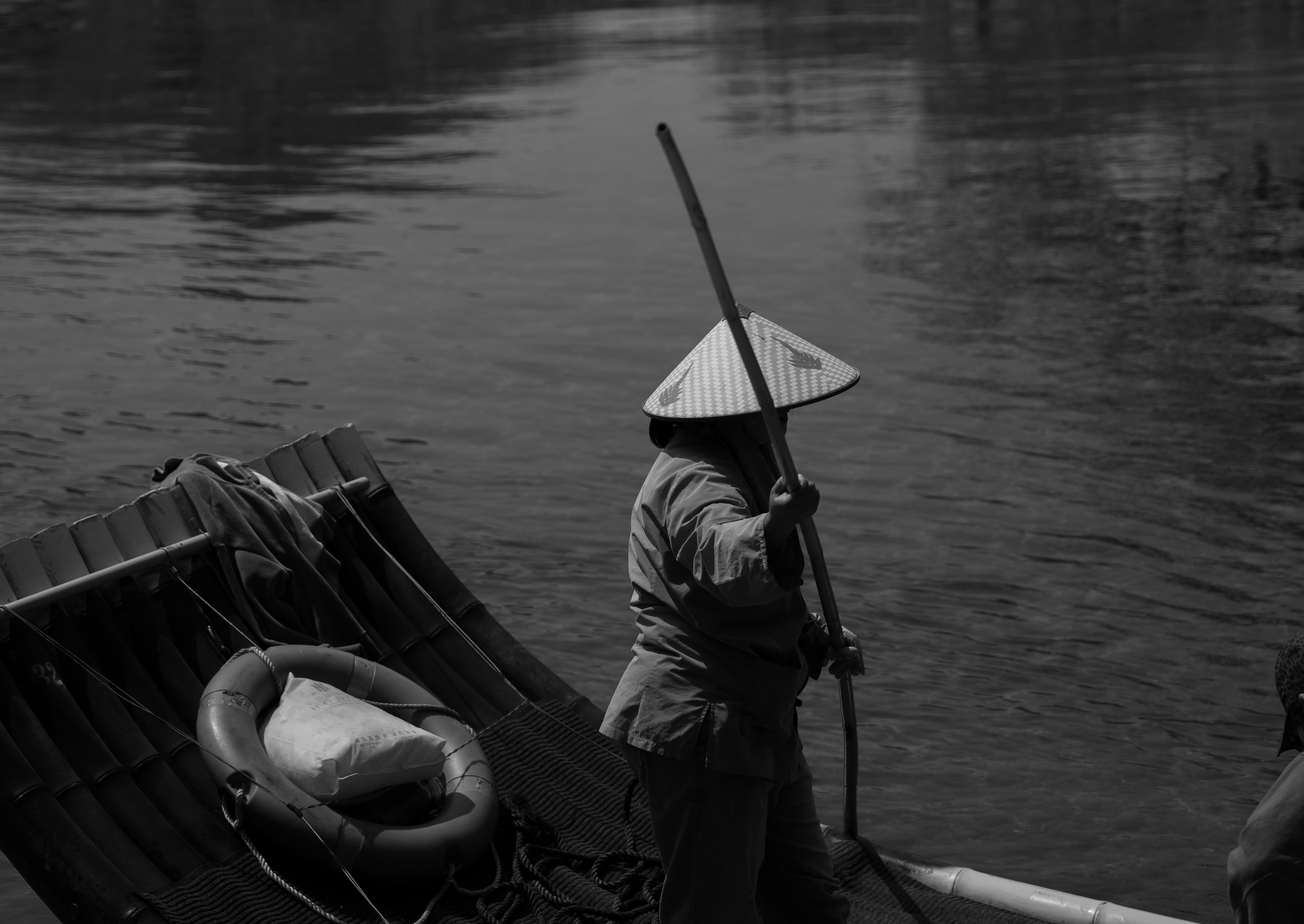 Person in traditional attire standing on a boat with a pole on a calm river.