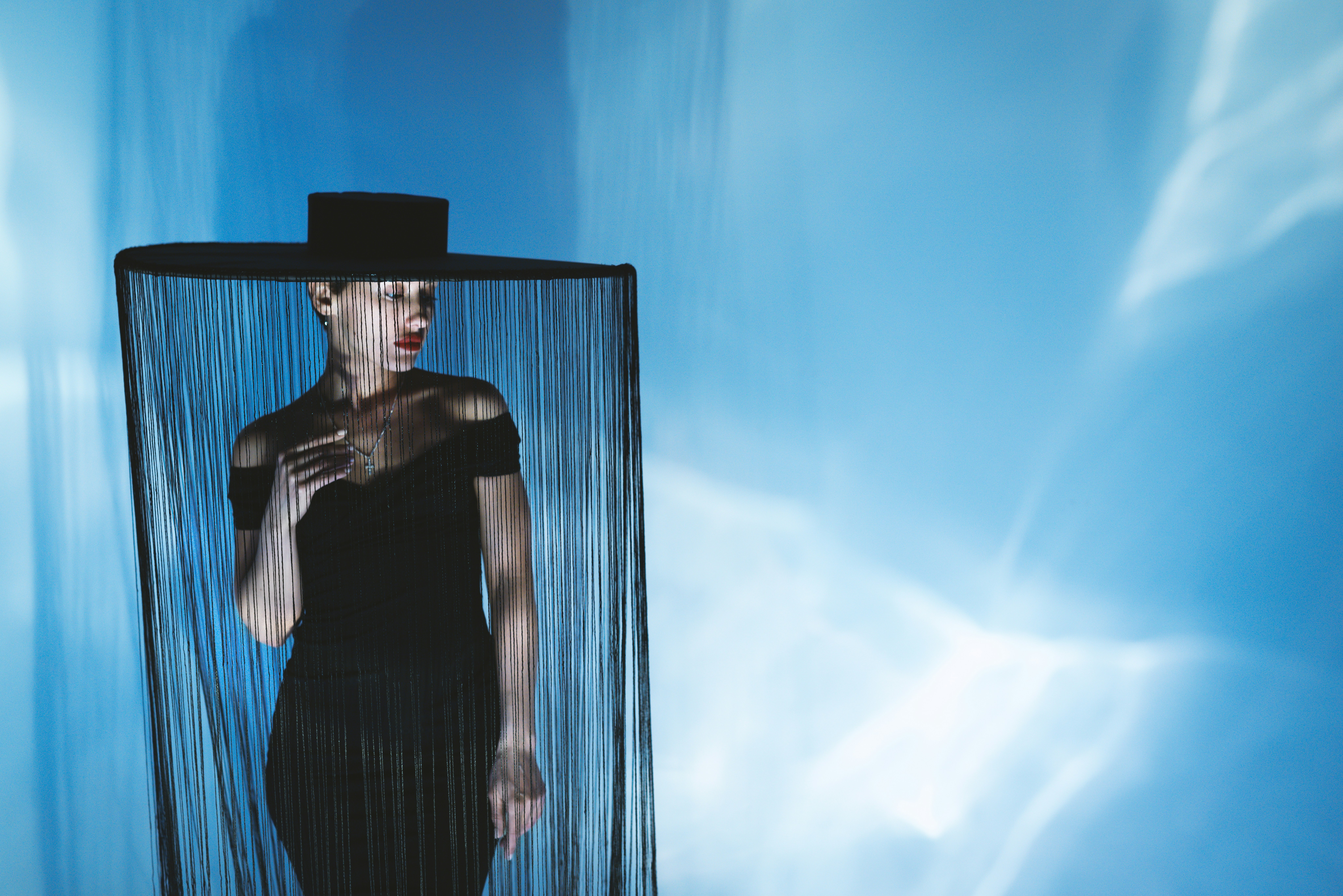 Woman in black dress and hat stands behind sheer black fabric against a soft blue background.
