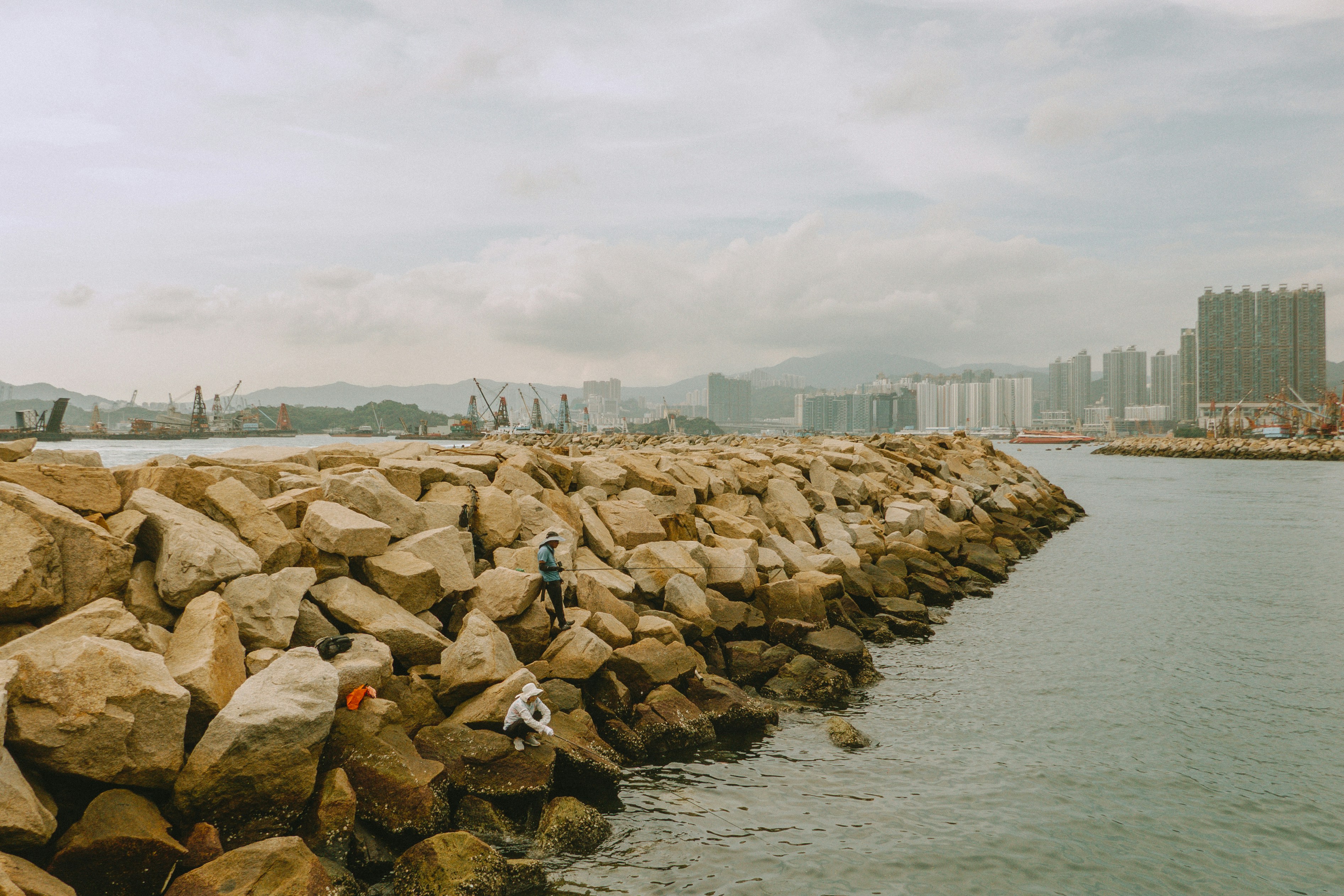Two fishermen cast lines among large boulders at a rocky shore, with barges and the Kowloon skyline in the background.