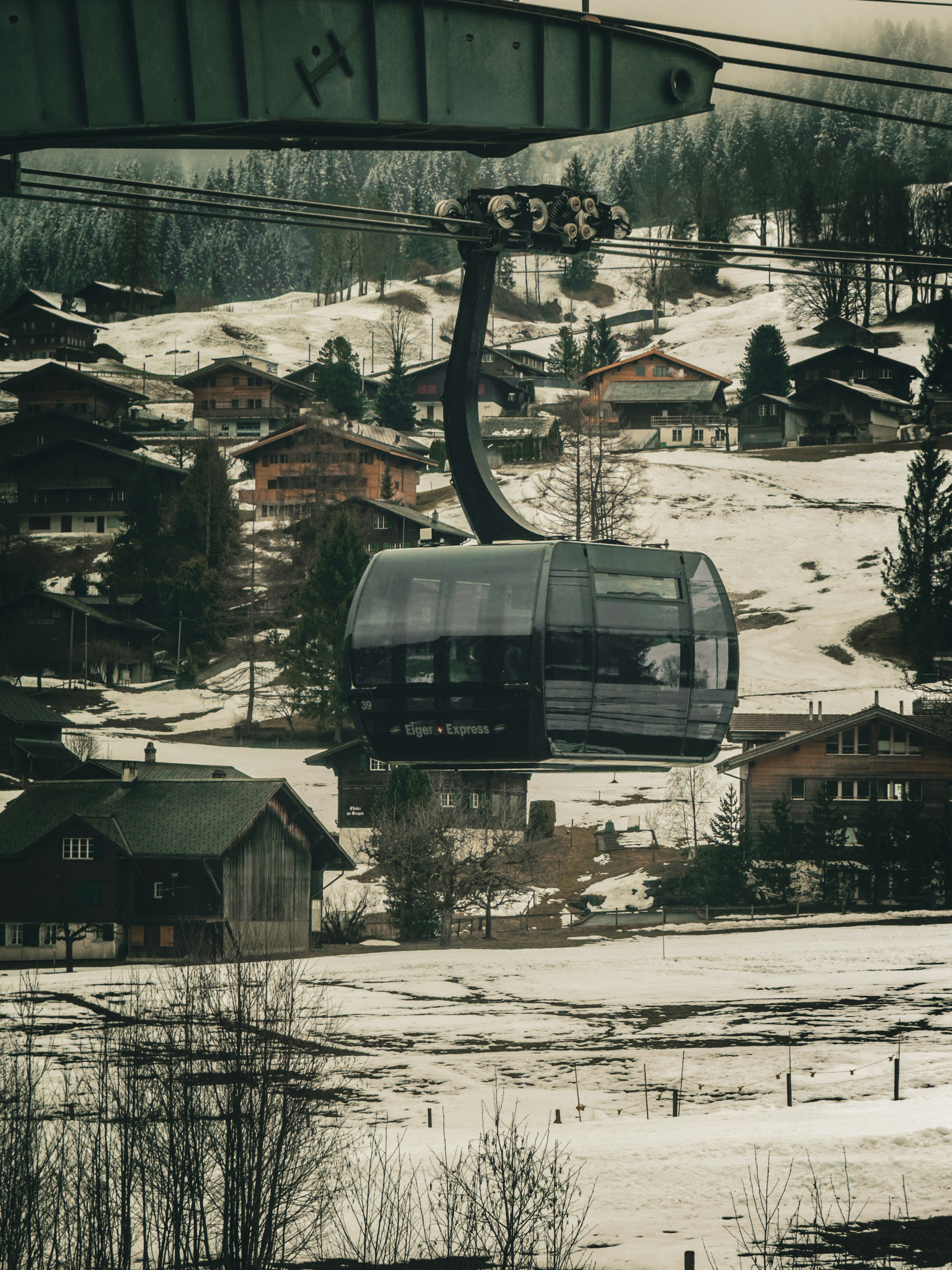 Cable car glides above a snow-dusted alpine village with rustic wooden houses.