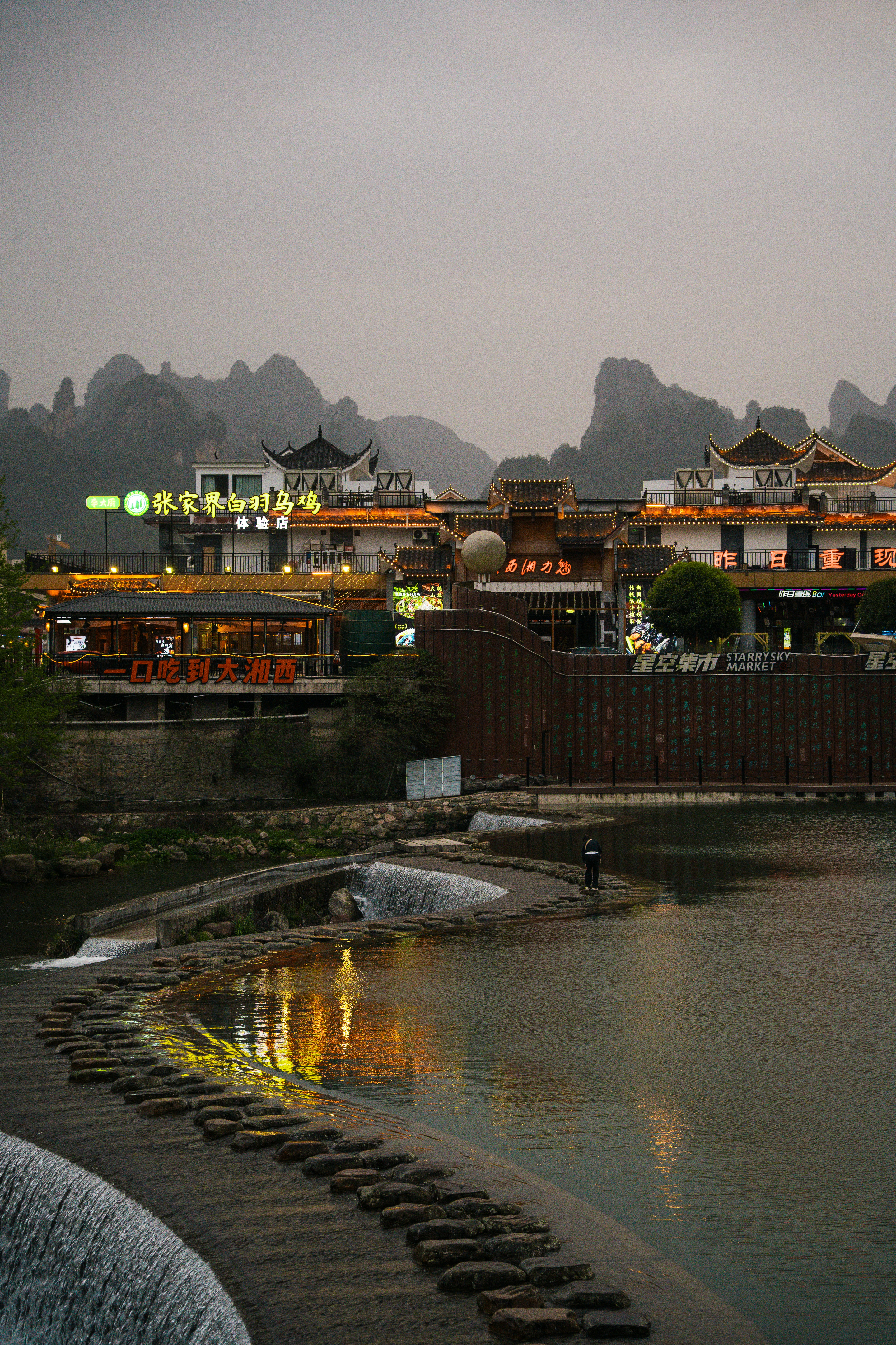 Neon-lit riverside bazaar beneath distant karst silhouettes