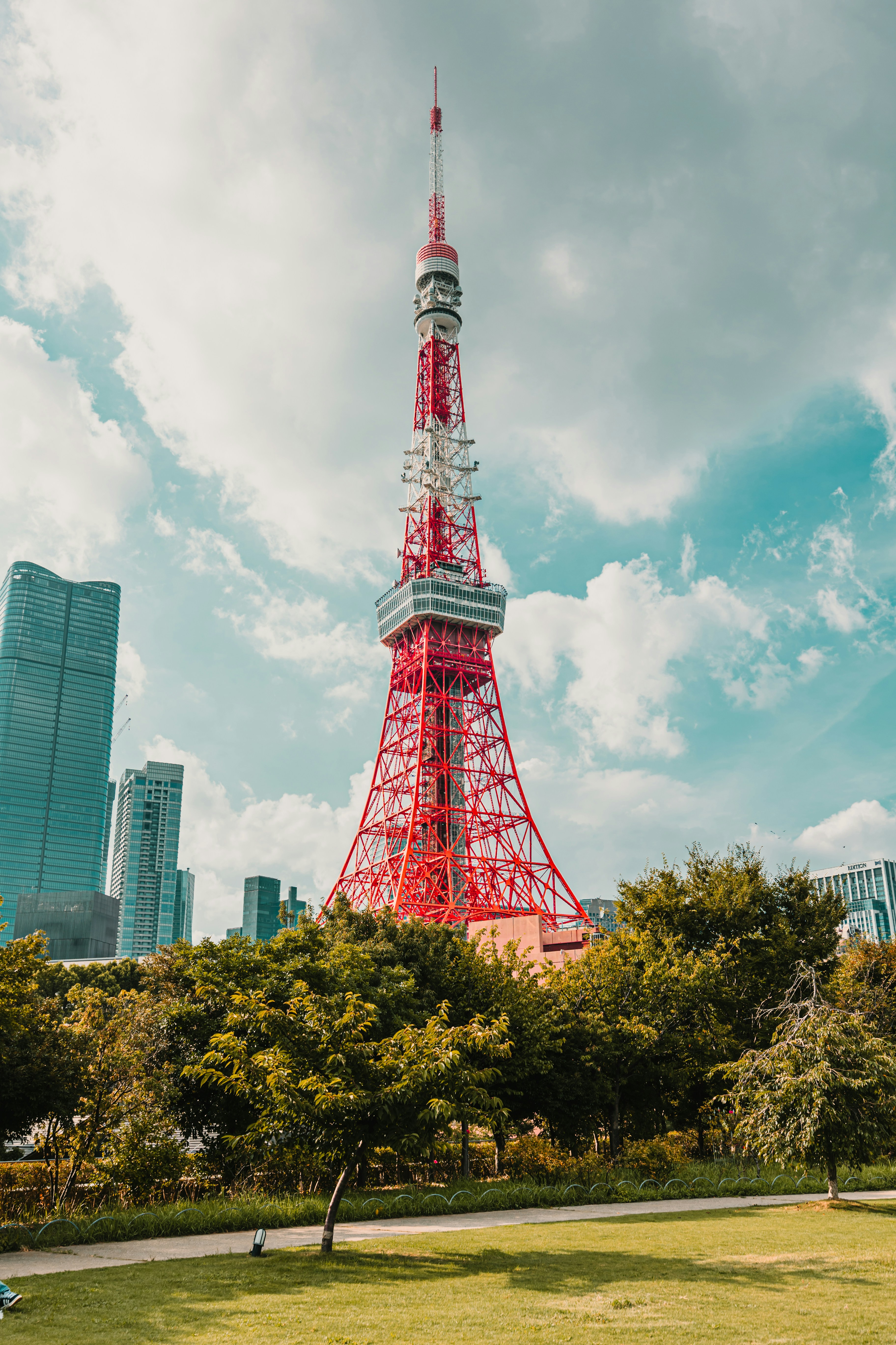 Tokyo Tower rising above green trees and a park, with modern skyscrapers in the background and a partly cloudy sky.
