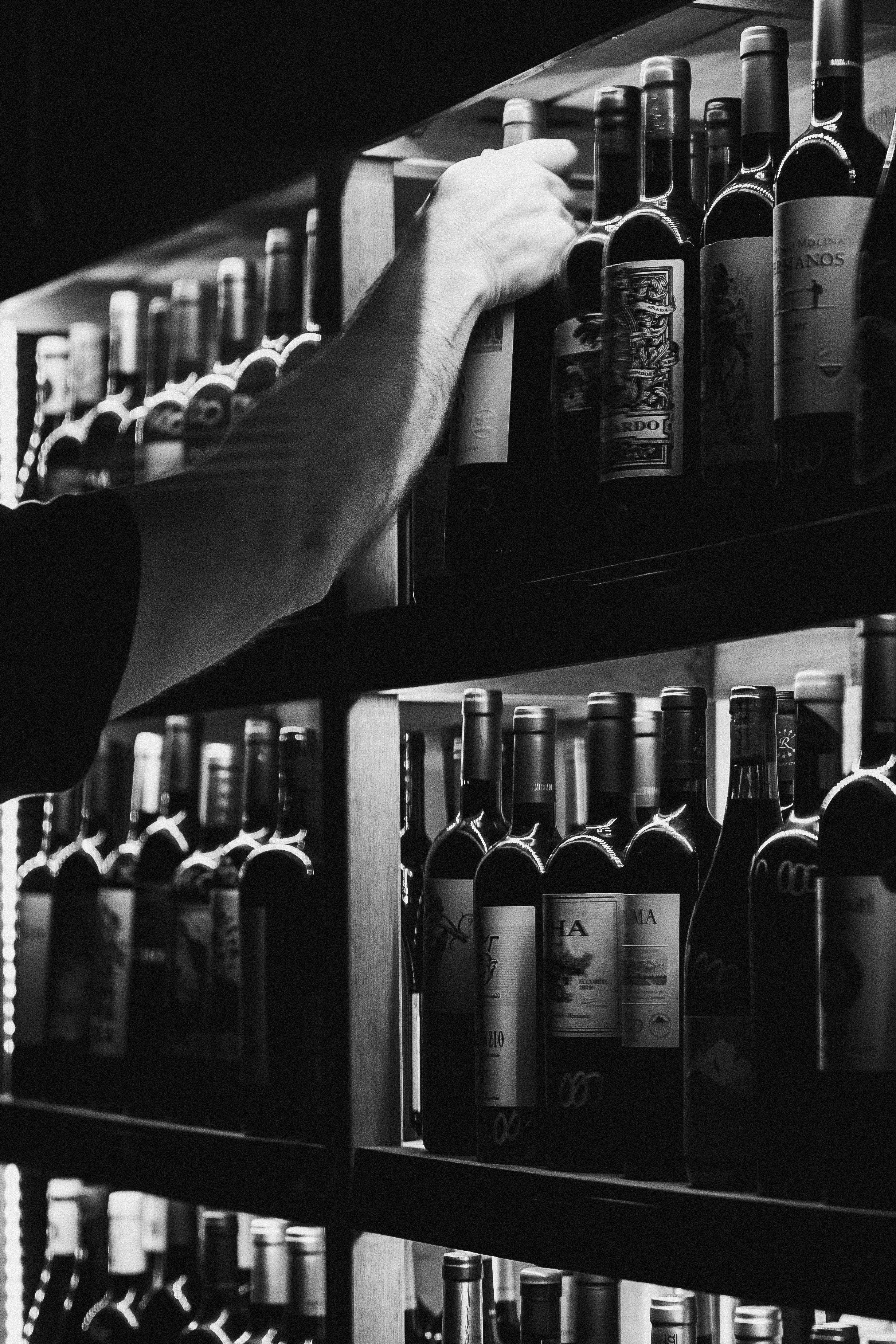 Black and white scene of a hand reaching for a wine bottle on a dimly lit shelf.