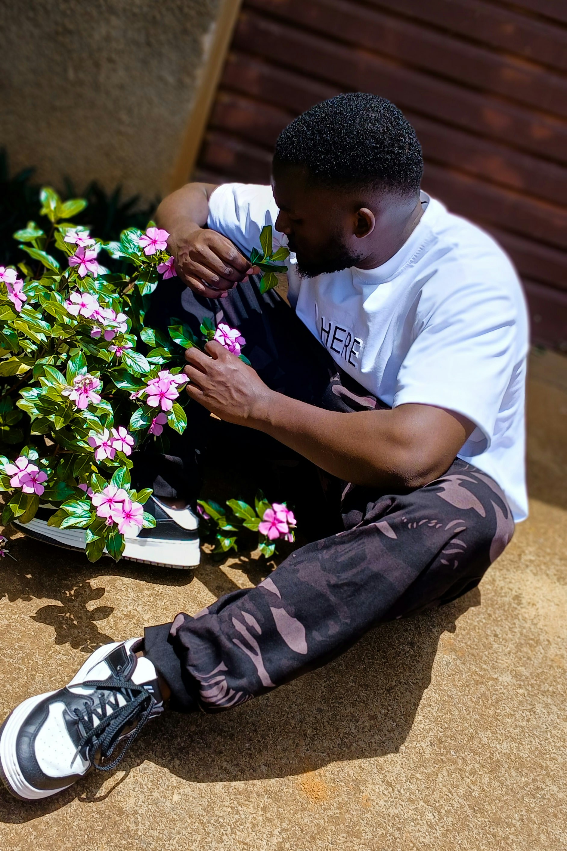 Candid photo of a man seated on a concrete step, closely inspecting pink blossoms beside a leafy plant.