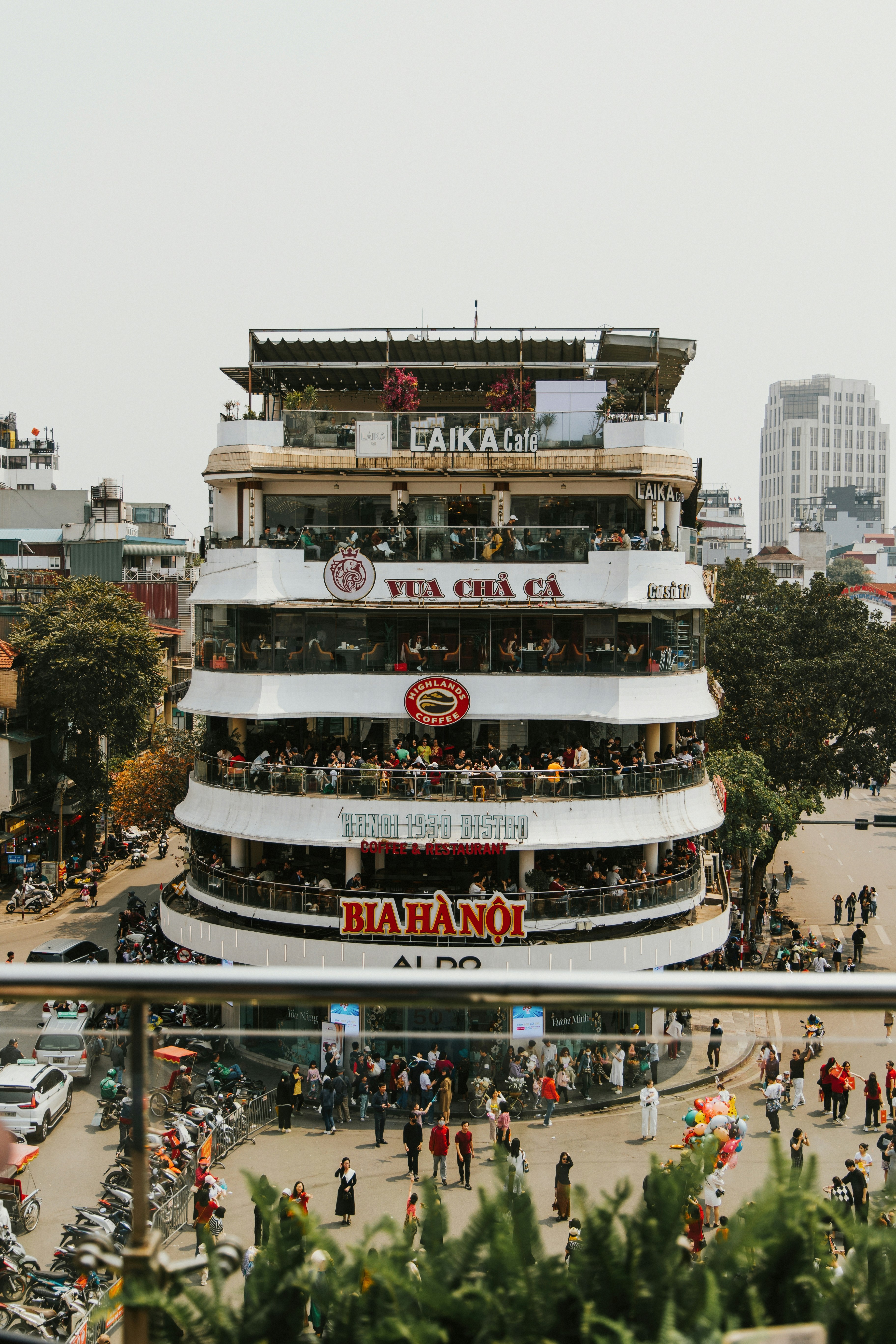 Multi-level café and restaurant bustling with patrons, showcasing lively street activity in a cityscape. The structure features various signage and greenery on the balcony.