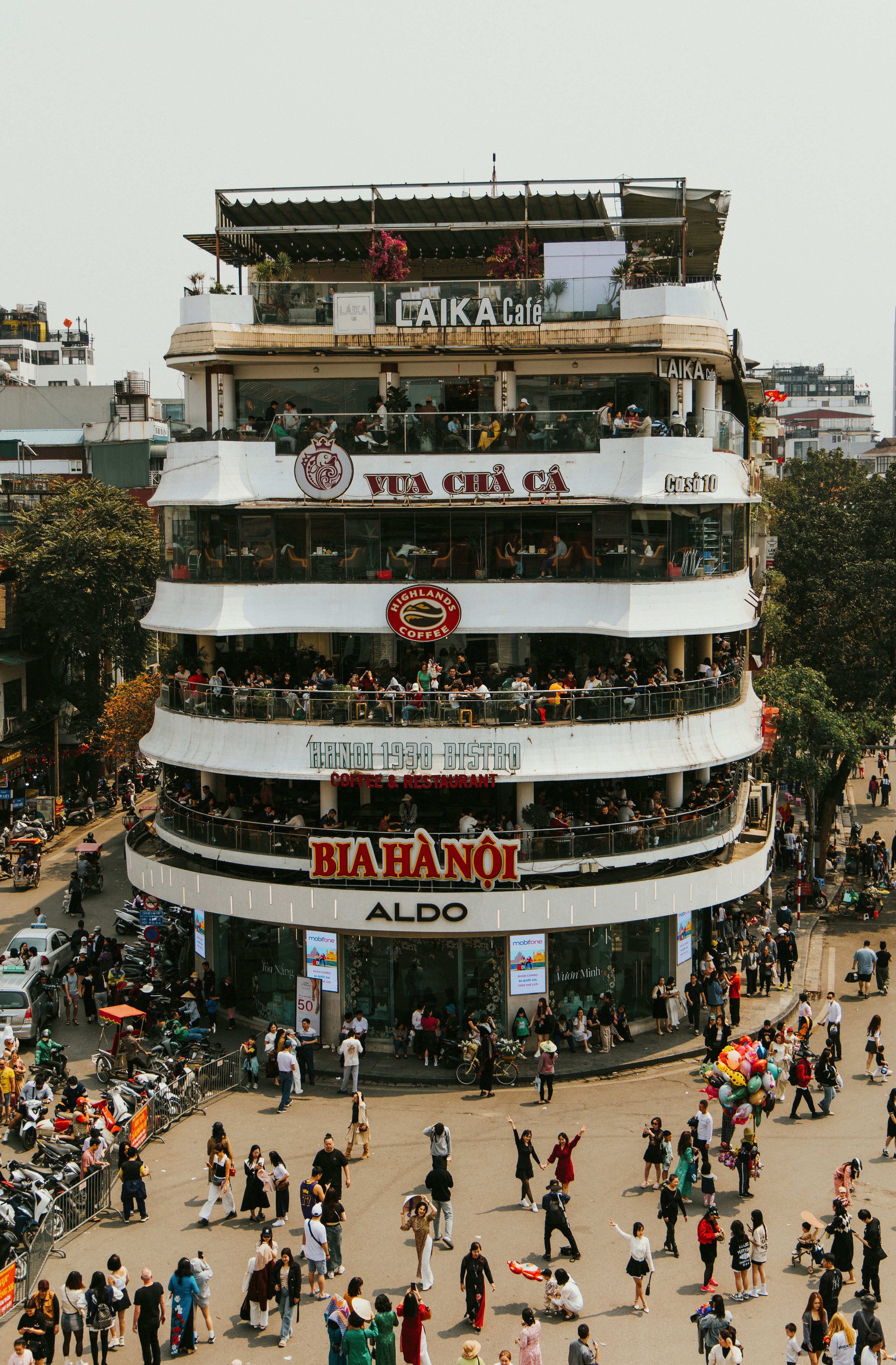 Multi-story café bustling with patrons, surrounded by vibrant street life and shops. The architecture showcases a blend of modern design and urban culture.