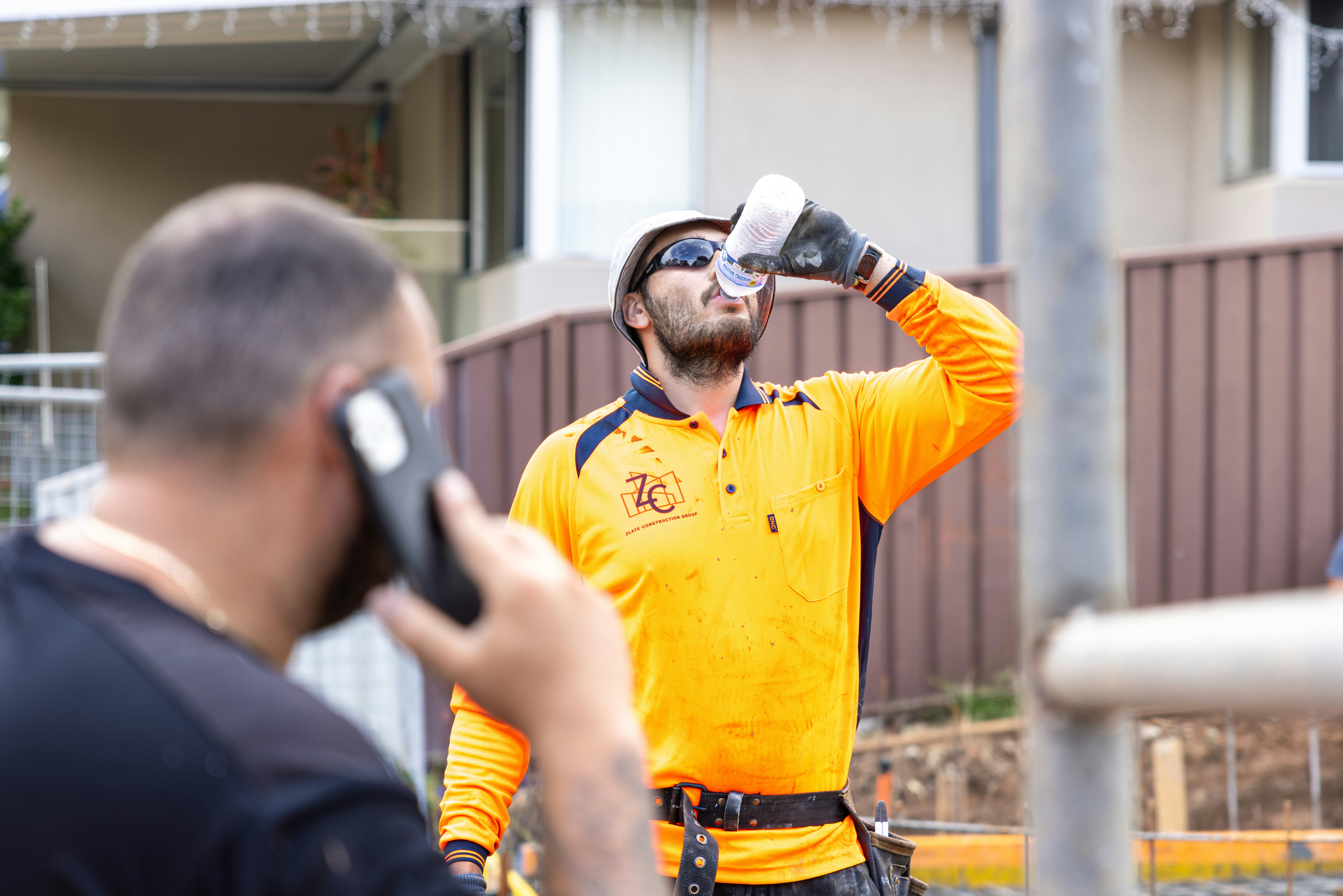 Construction worker in orange attire takes a drink from a bottle while another uses a phone in the foreground.
