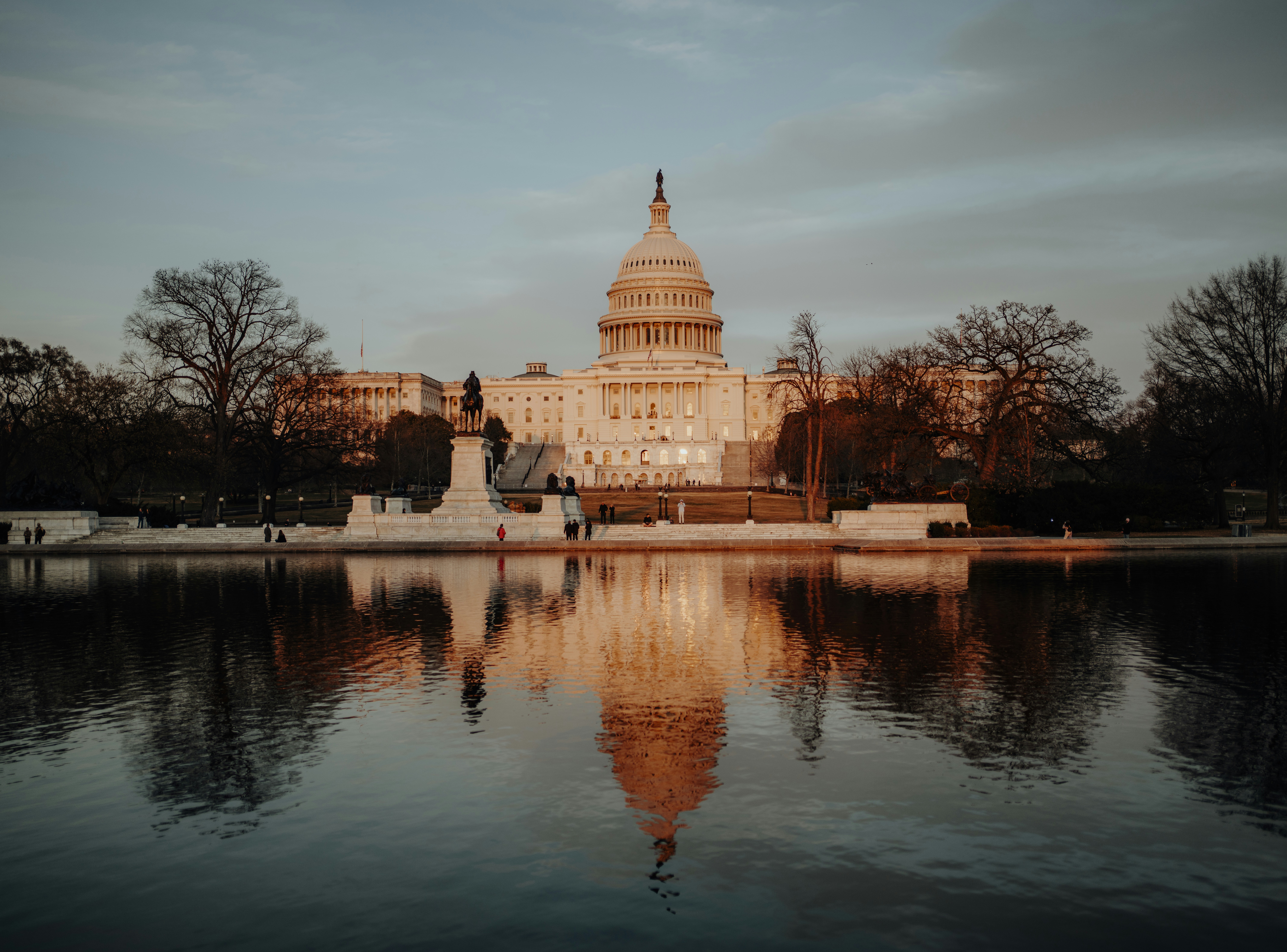 The U.S. Capitol building reflected in a tranquil pool at sunset, with a warm glow illuminating the scene.