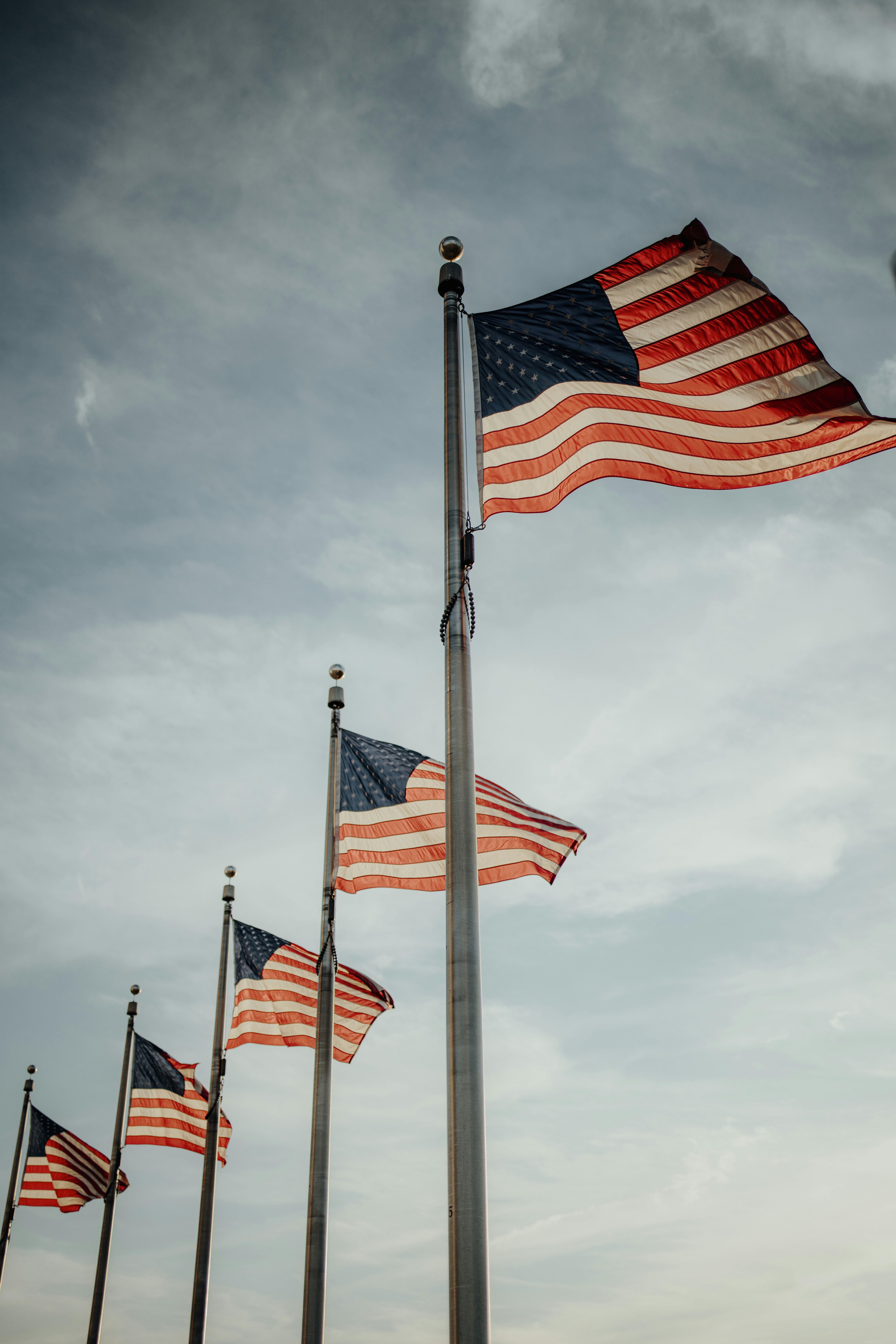 Multiple American flags fluttering on poles against a backdrop of a cloudy sky.