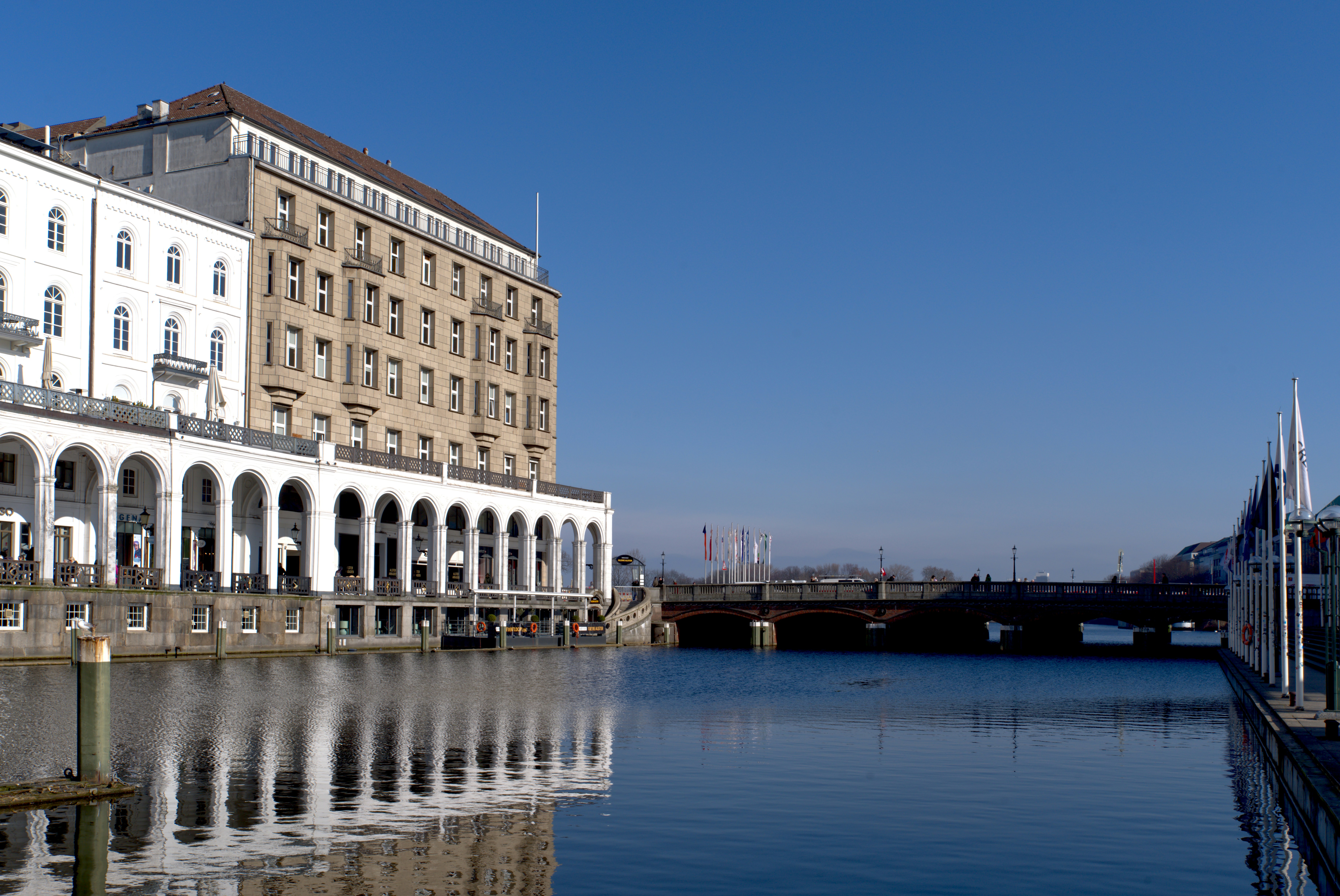 Historic building with arched colonnade reflected in calm waters under a clear blue sky.