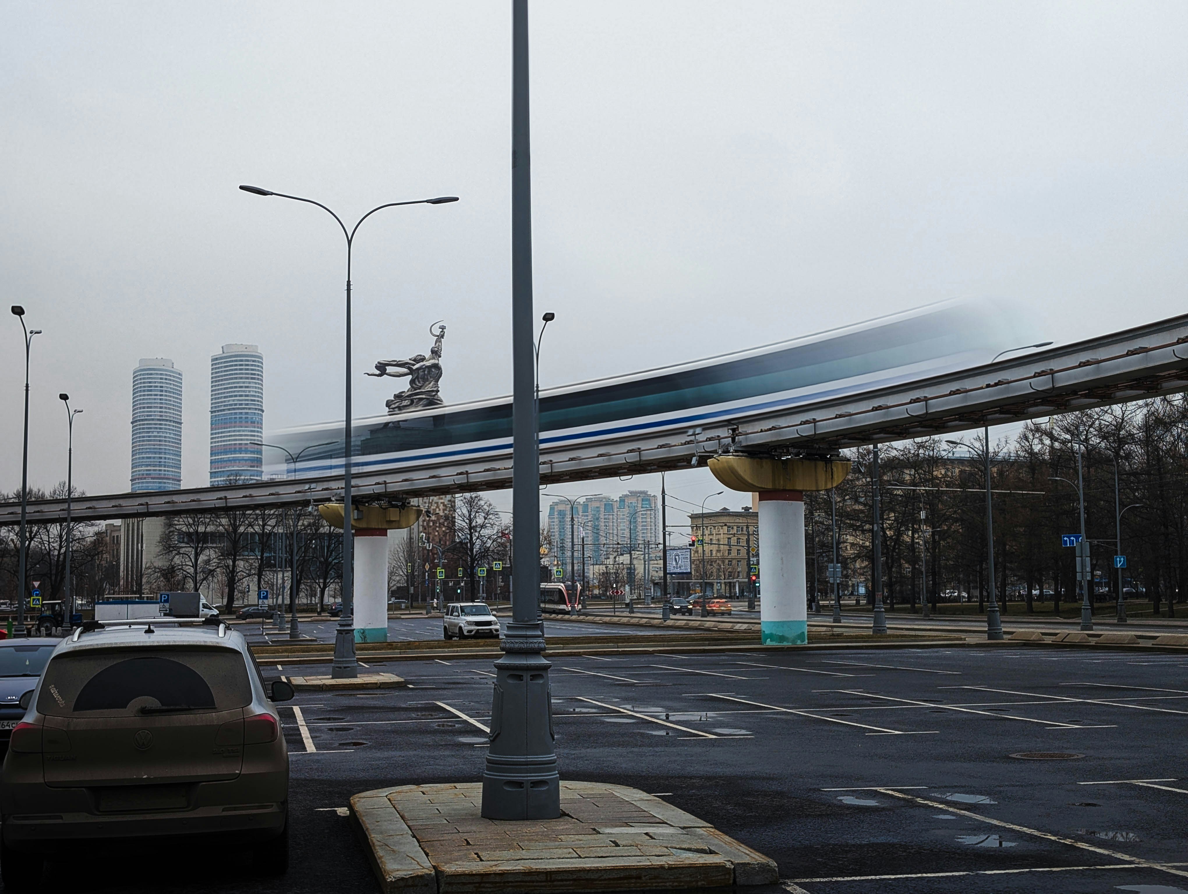 Motion-blurred elevated train crosses a city parking lot, with a statue atop the rail supports beneath a gray, overcast sky.