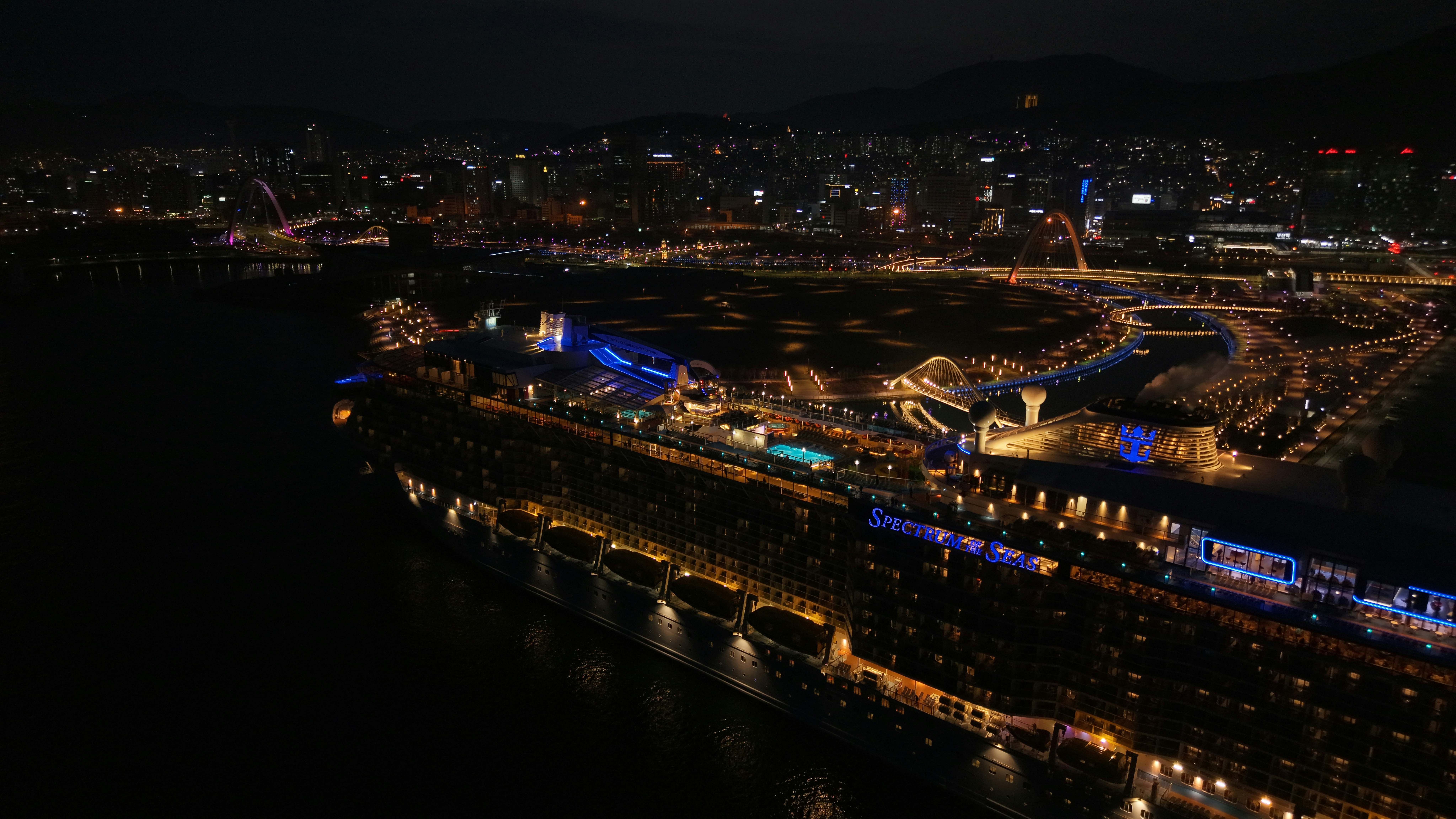 Illuminated cruise ship docked at Busan port with city skyline glowing at night.